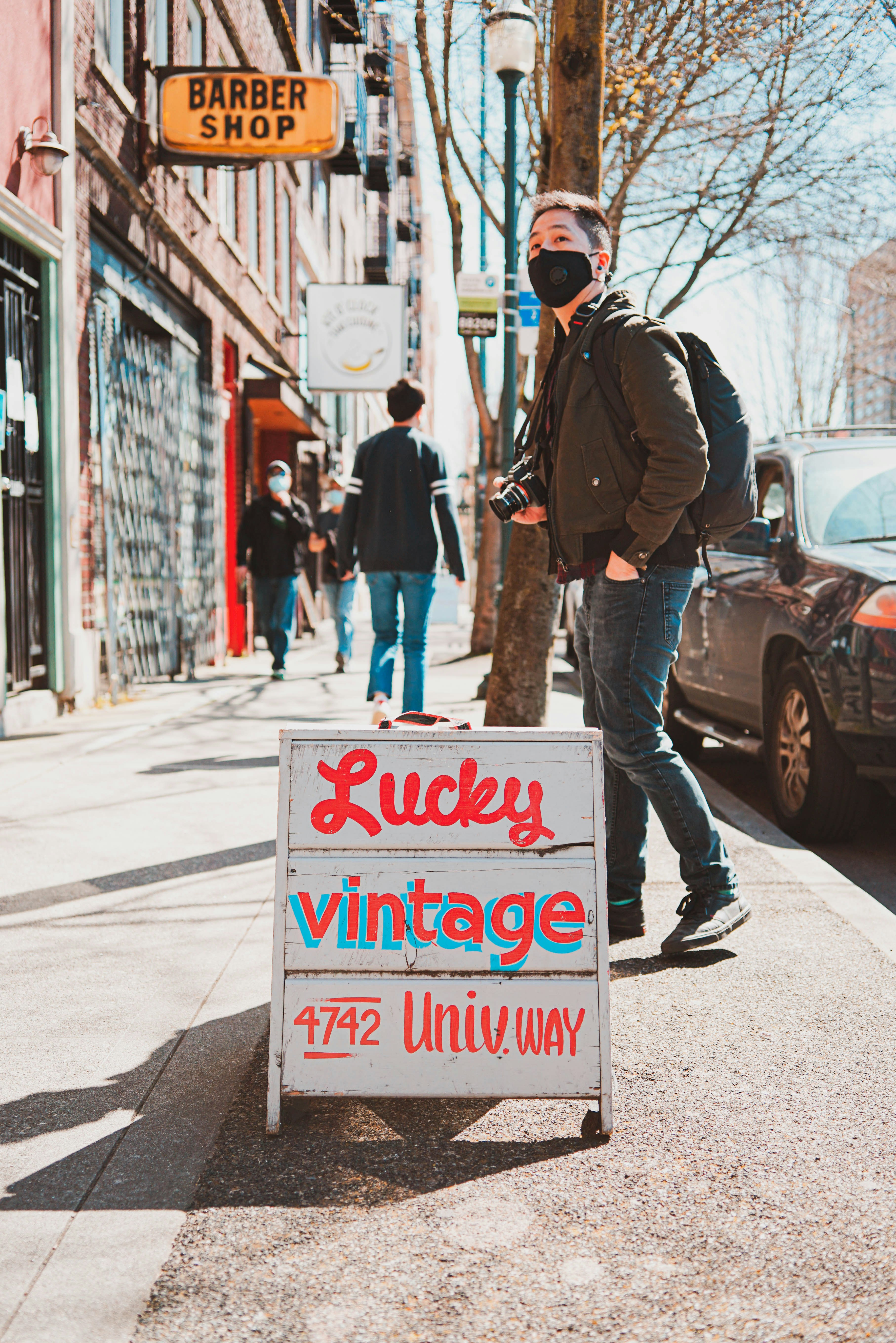 Mask, Camera, and Street: A Candid Pause Outside a Vintage Sign