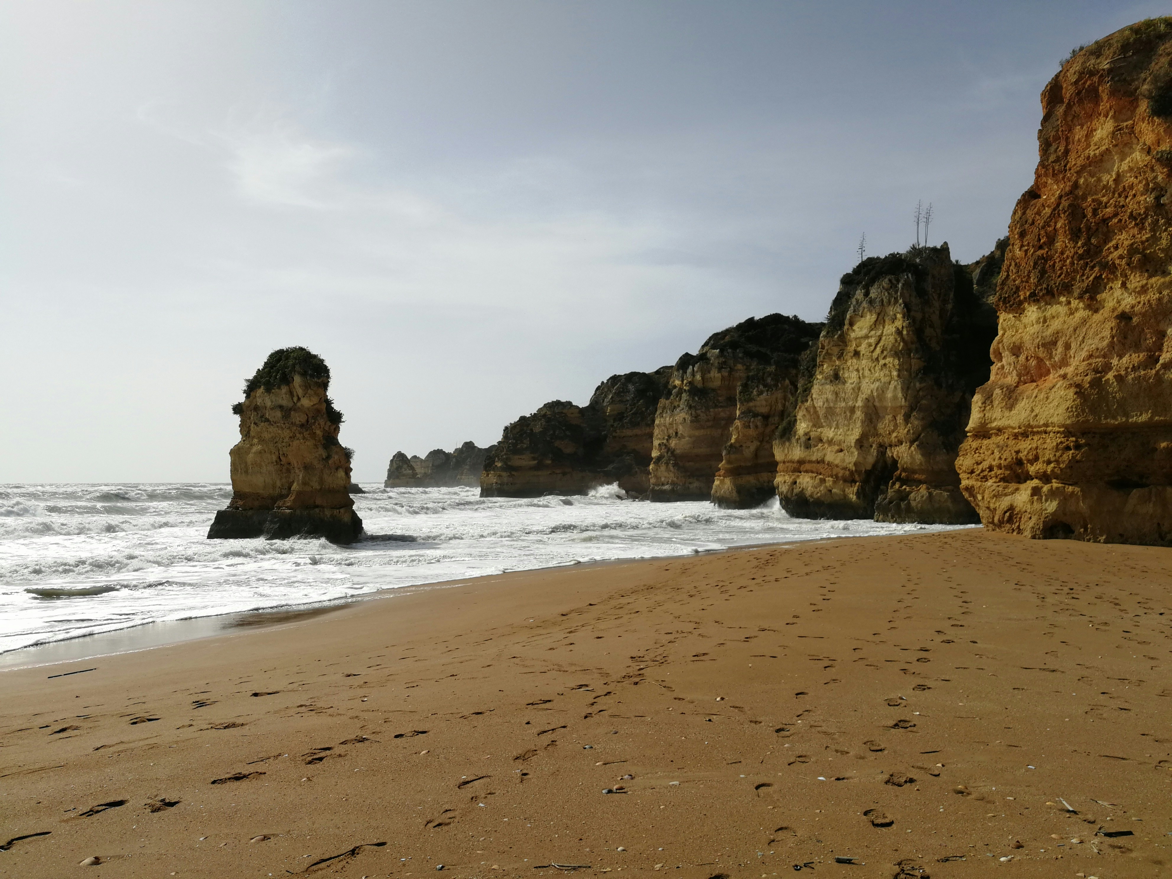 Towering brown rock formations line a sandy shore with waves gently lapping the coast under a clear sky.