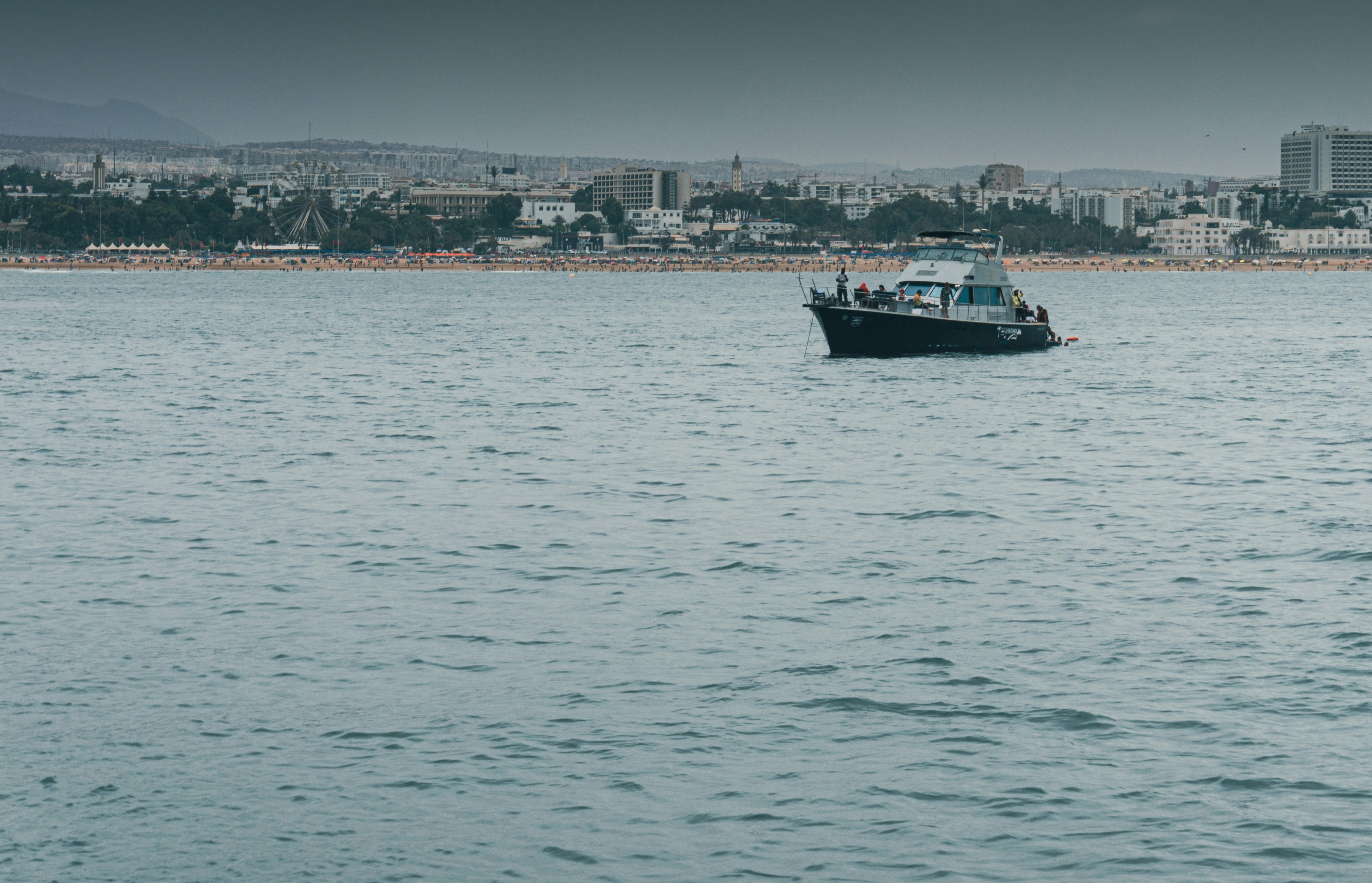 A lone boat sails on calm waters with a distant cityscape under an overcast sky.