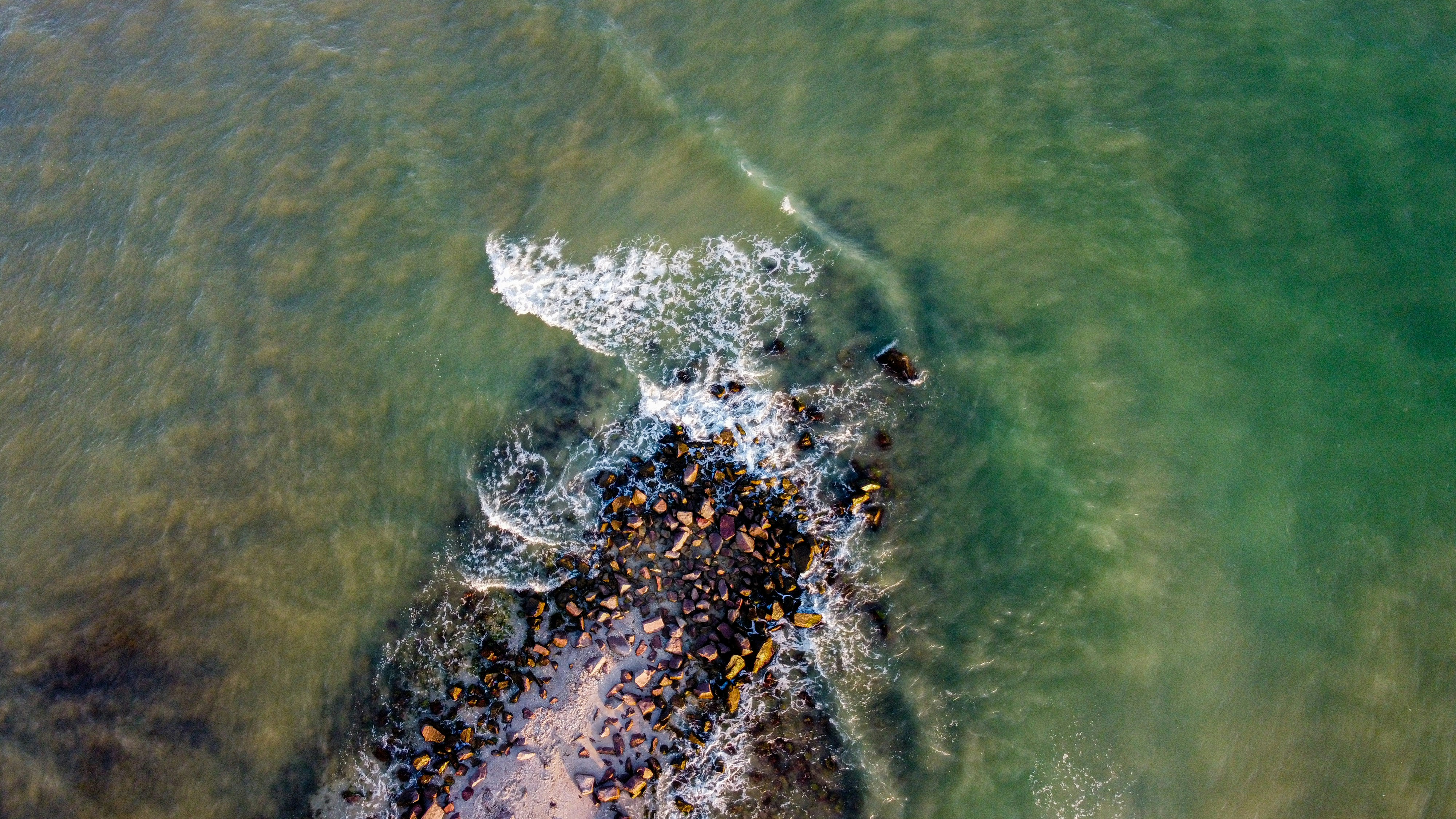 Aerial view of rocky shoreline meeting gentle waves, showcasing the interplay of land and sea. The vibrant colors of the water contrast with the rugged textures of the rocks.