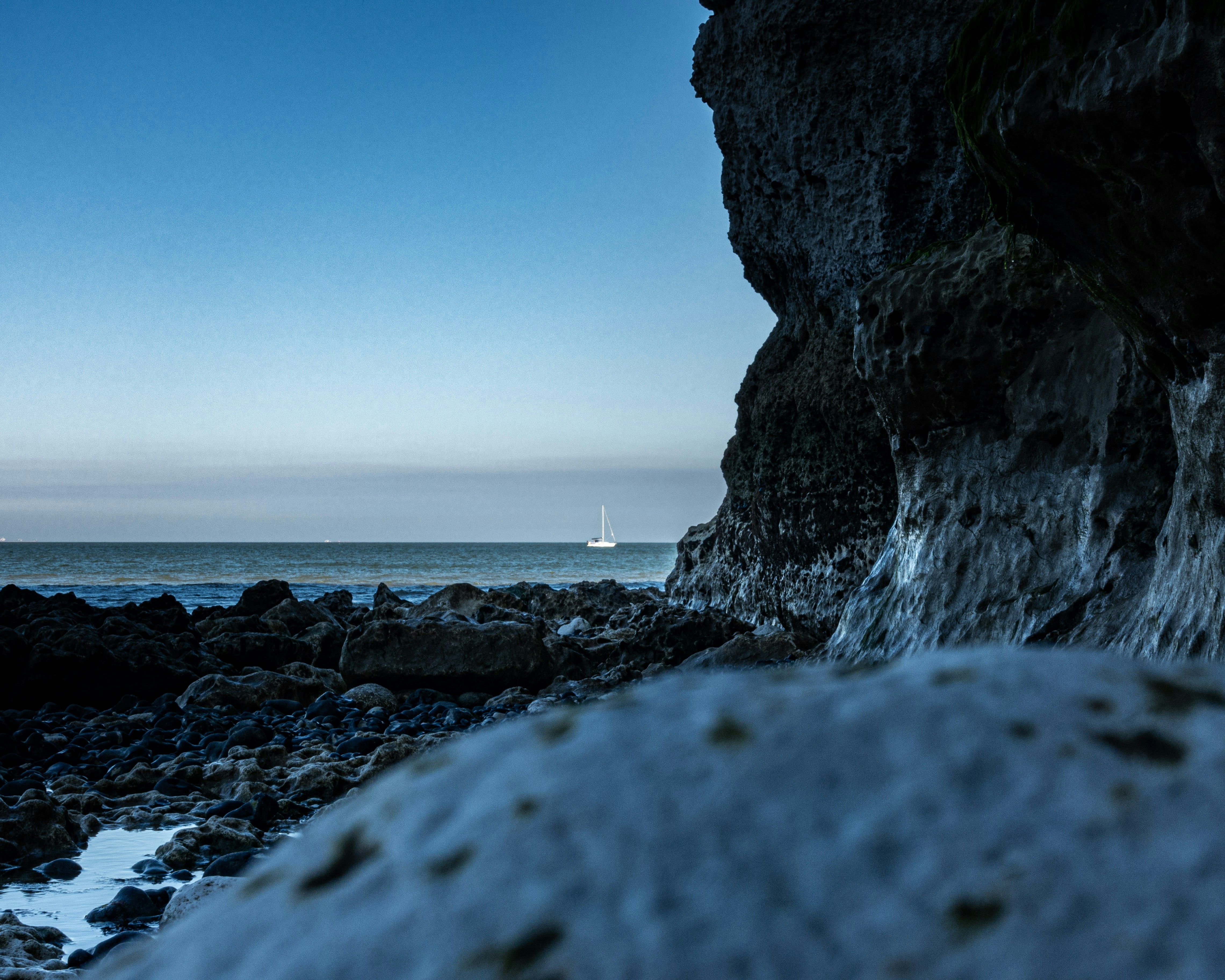 Rocky cliffside overlooking a calm sea with a small sailboat on the horizon at dusk.