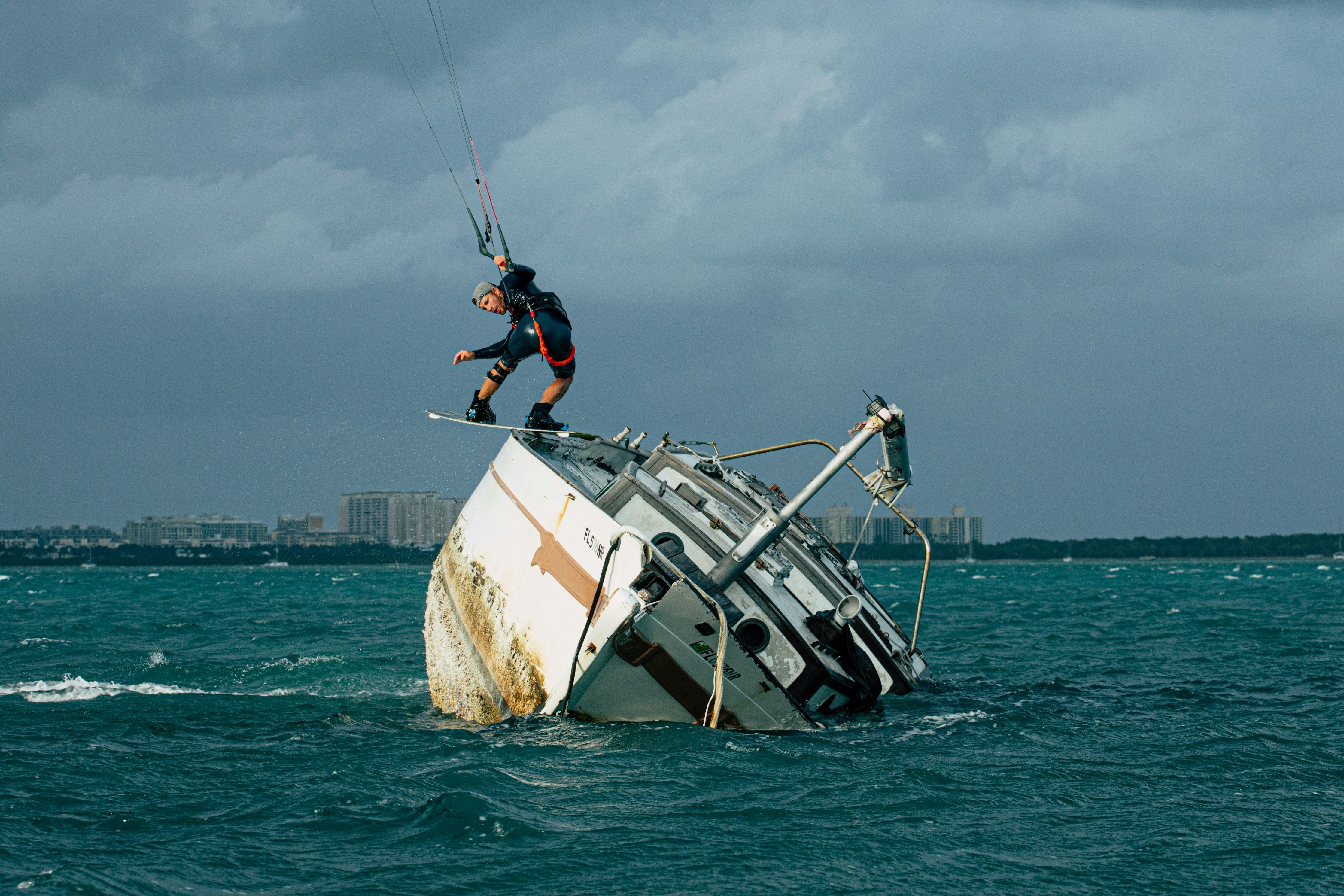 Kiteboarder performing a jump off the edge of a capsized boat in choppy waters, with a stormy sky in the background.