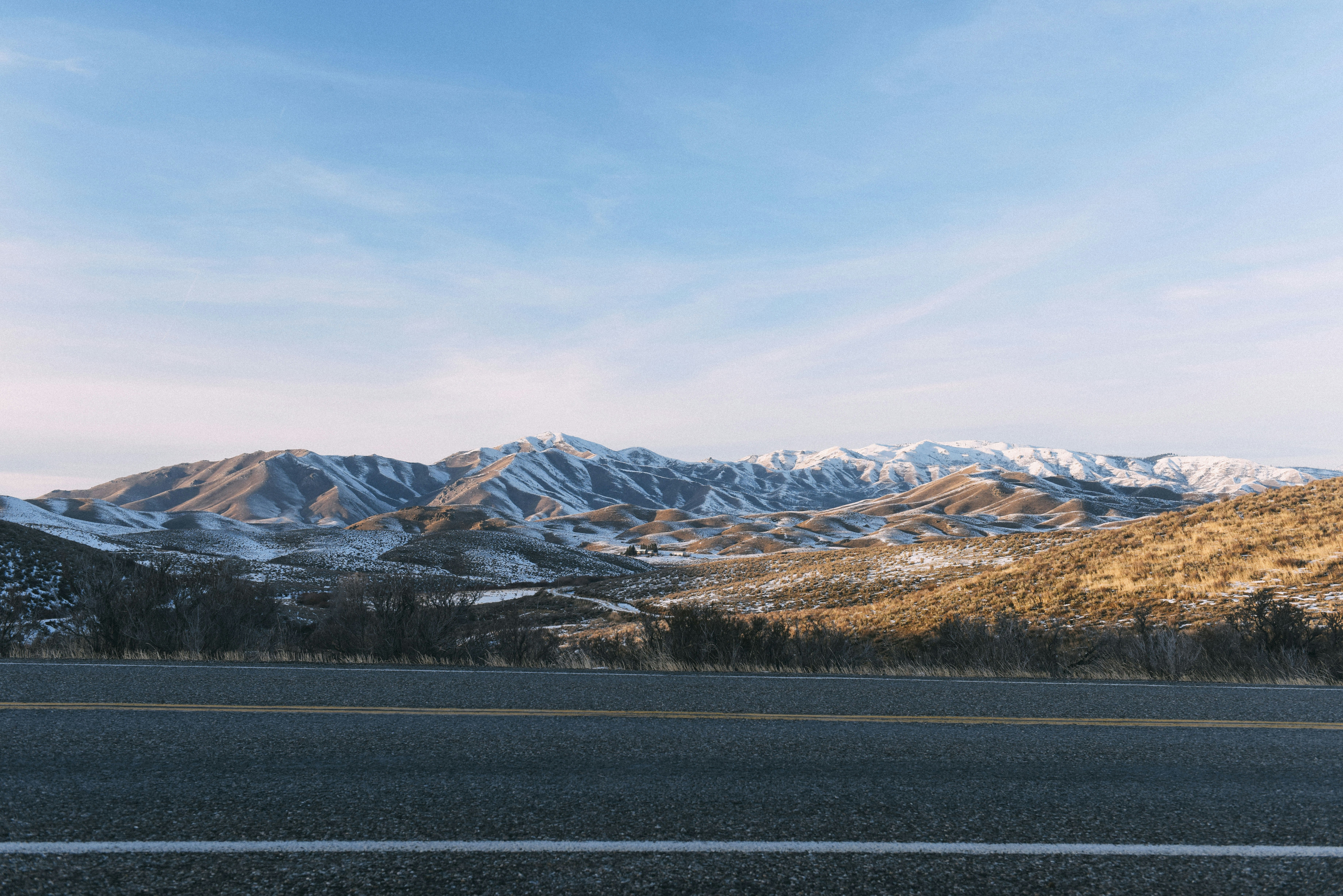 Rolling snow-capped mountains stretch across the horizon under a soft blue sky, with a winding road in the foreground.