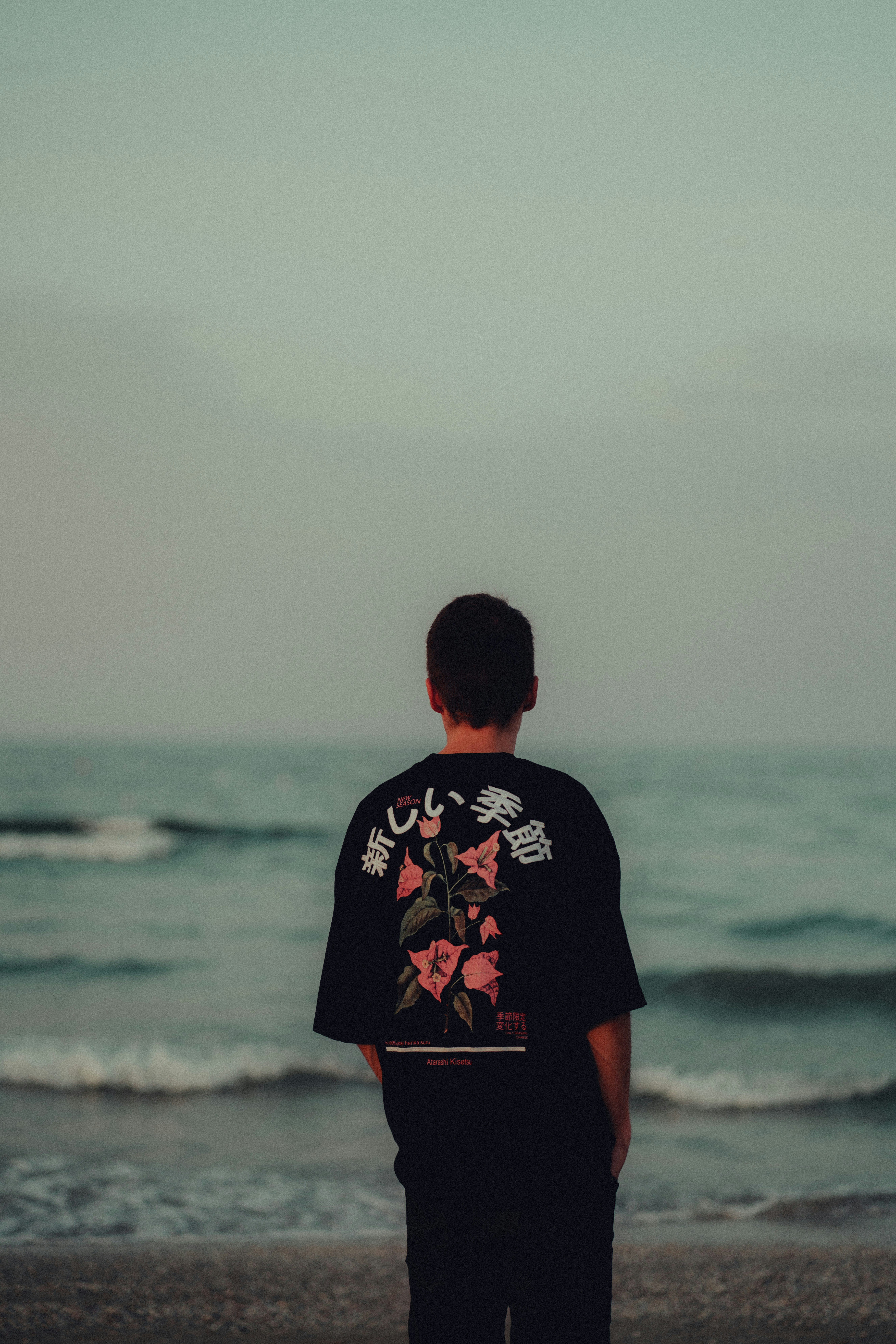 Silhouette of a person gazing at the tranquil sea, adorned in a floral graphic tee. The soft waves create a serene backdrop.