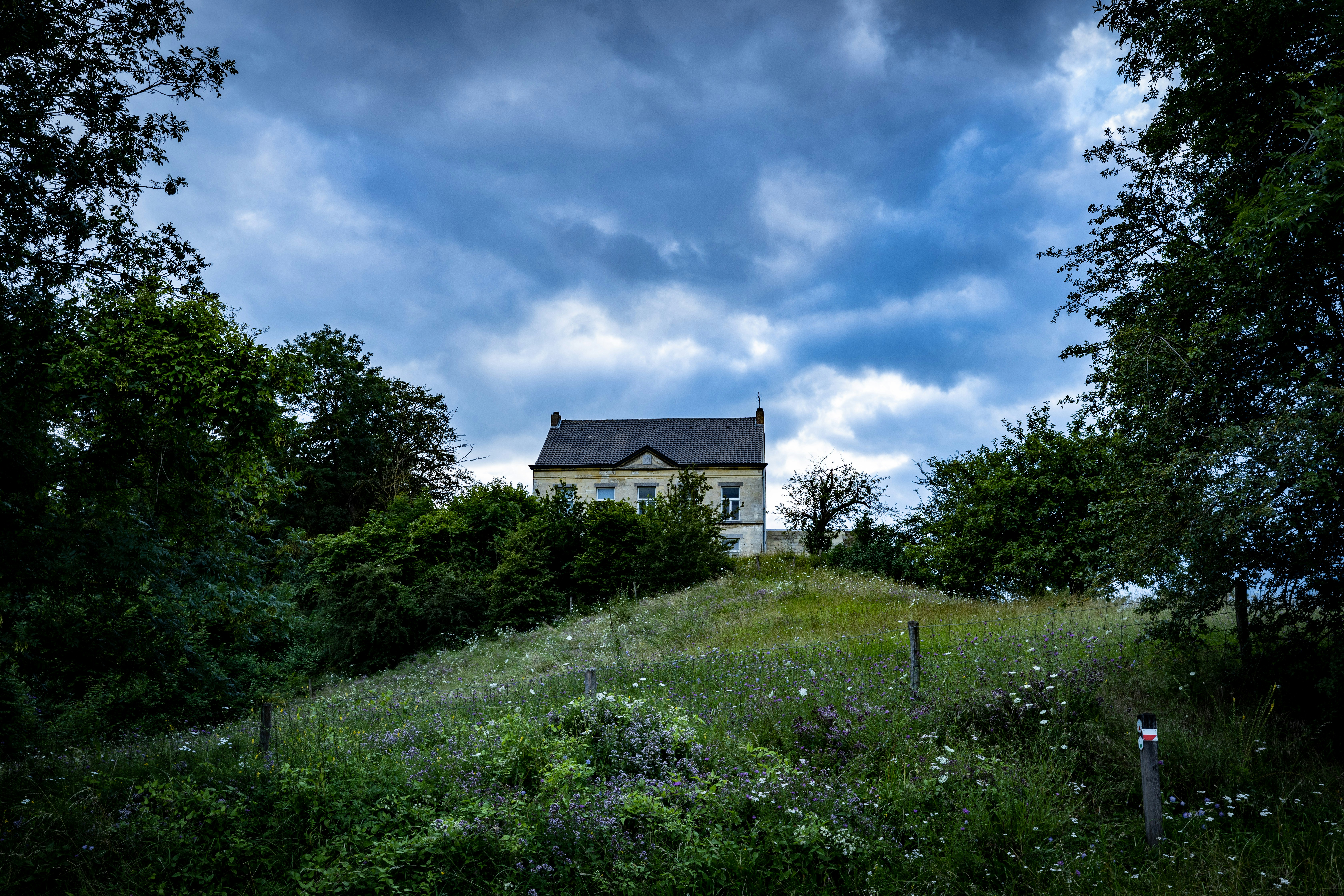 Historic house perched on a grassy hill, framed by trees under a dramatic cloudy sky.