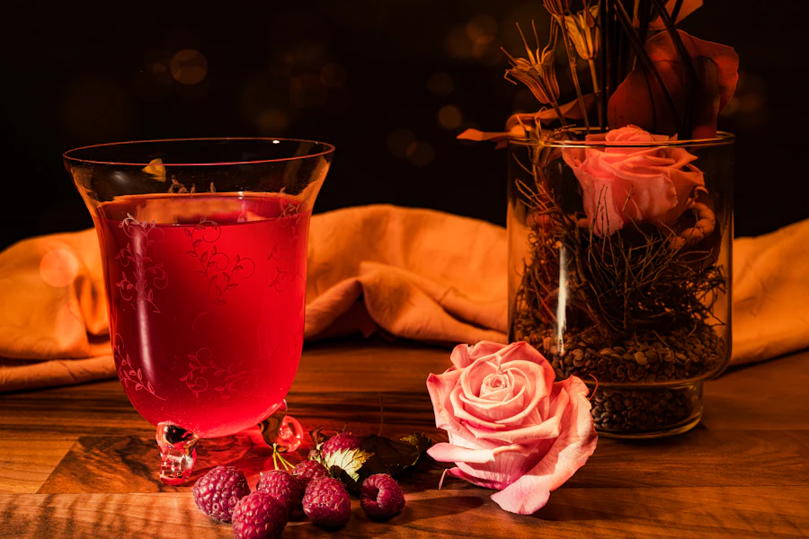 A red cocktail in a clear glass sits beside raspberries and pink roses on a wooden table, with a warm floral arrangement in the background.