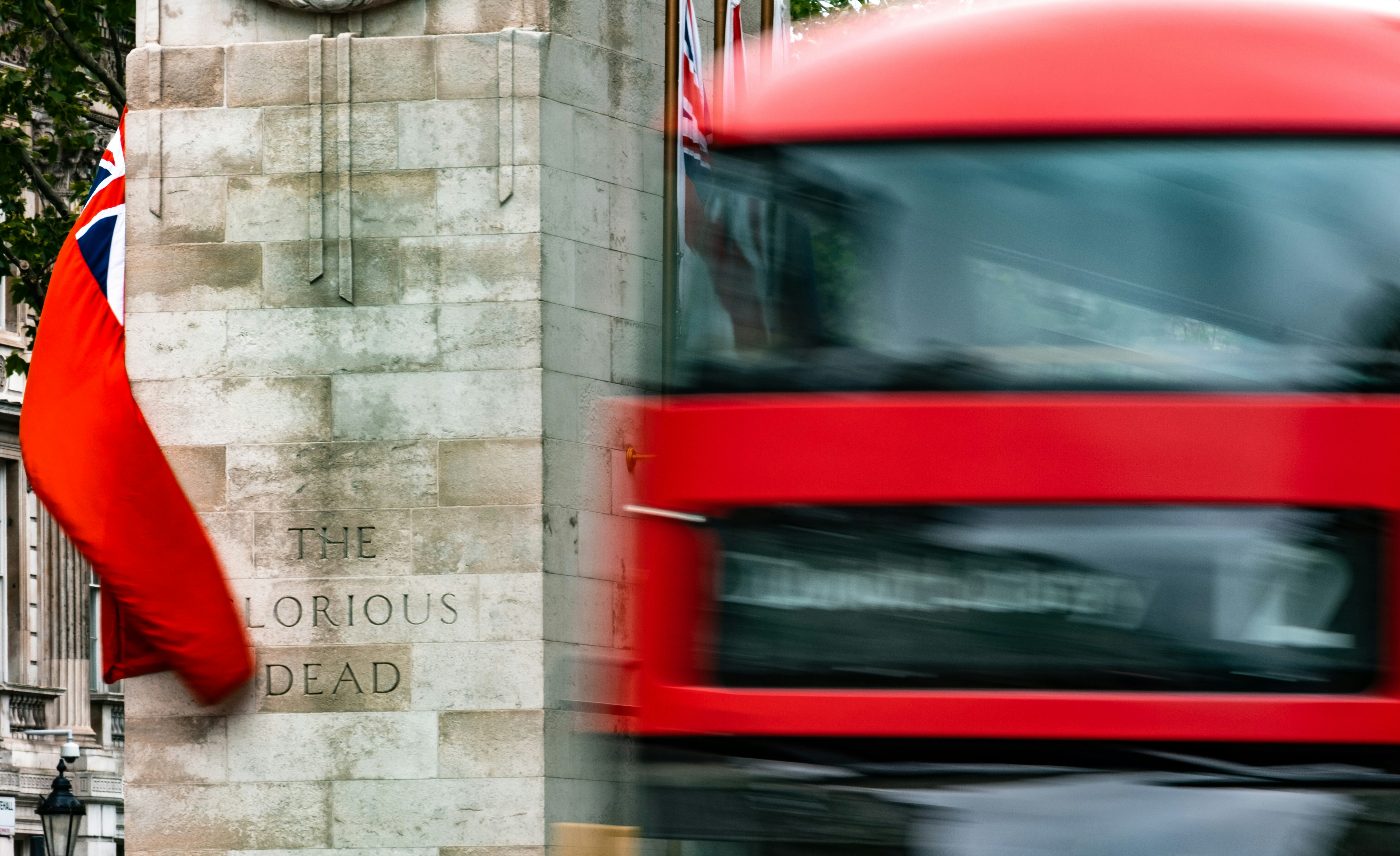 A red double-decker bus blurs past a stone memorial inscribed with 'THE GLORIOUS DEAD,' adorned with flags, symbolizing remembrance amidst urban life.