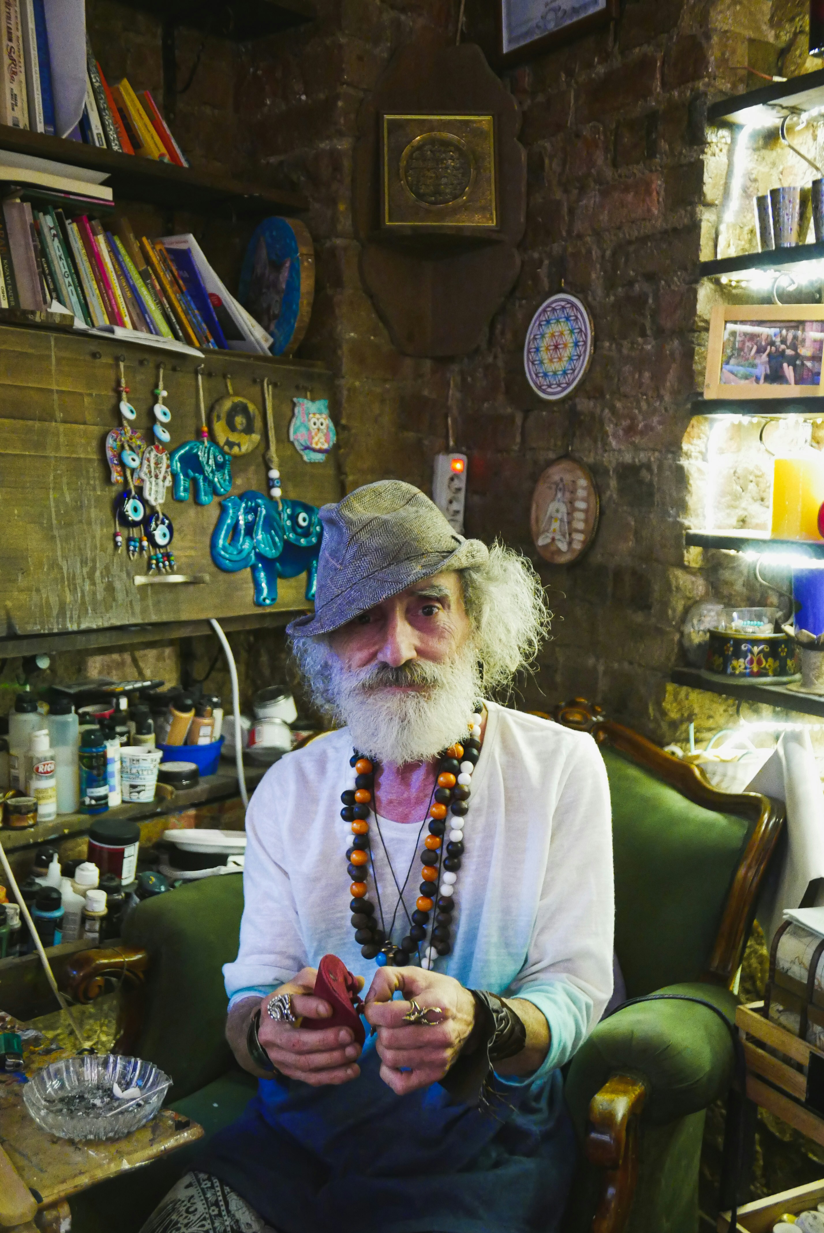 Bearded man with a hat seated in a cluttered workshop surrounded by books and trinkets.