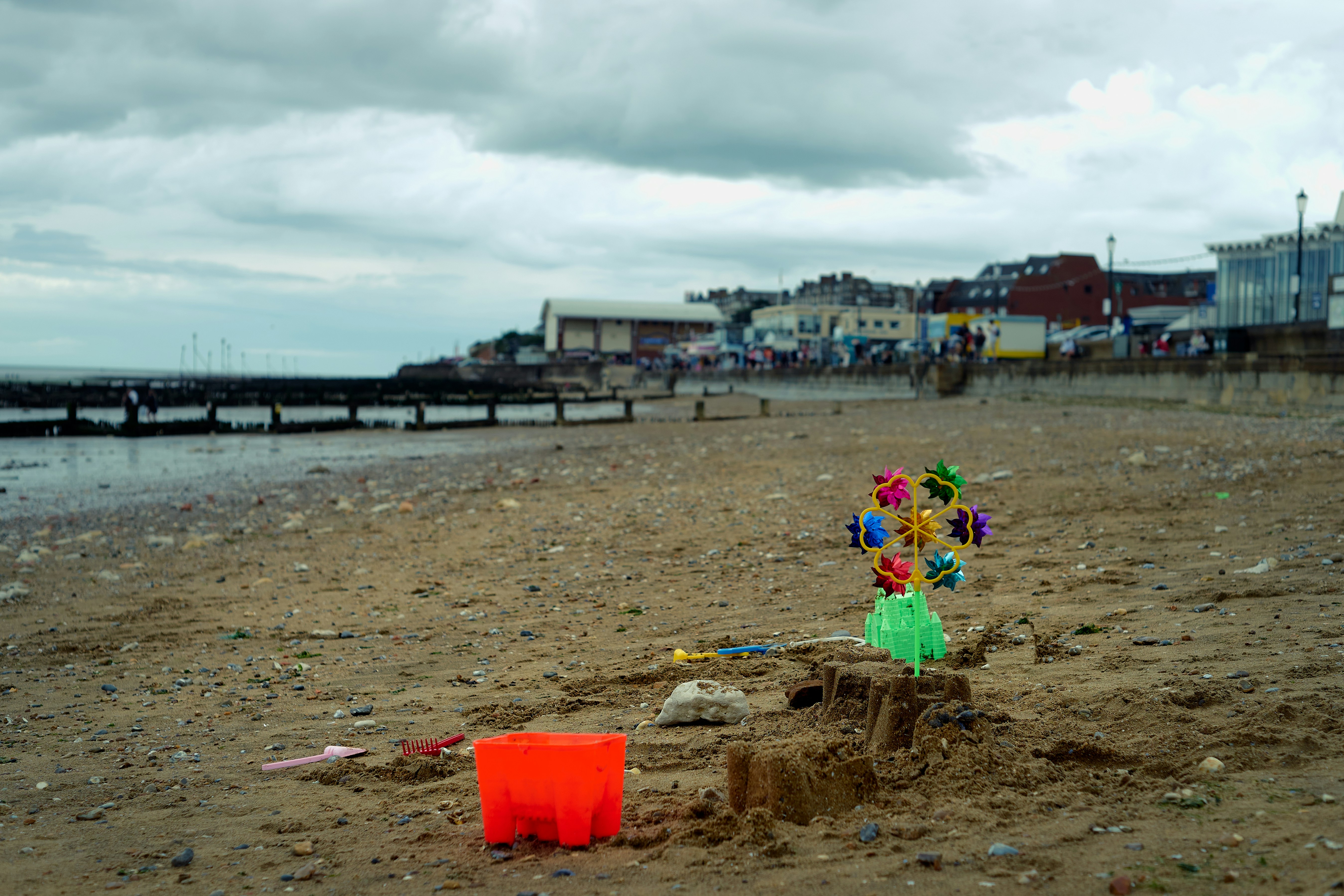 Colorful beach toys and a sandcastle sit on a sandy shore, with a cloudy seaside town in the background.