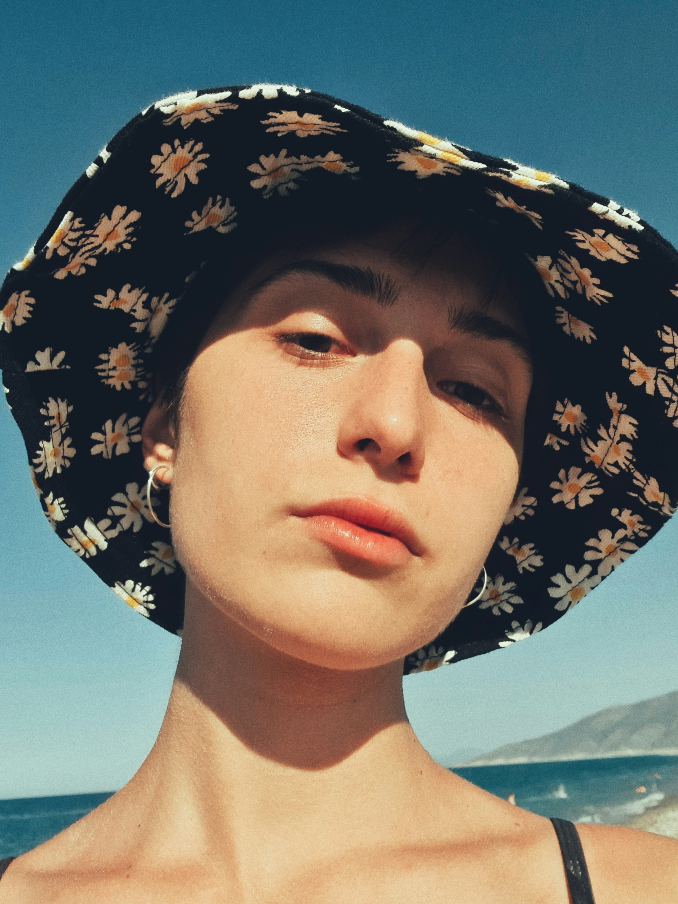 Close-up portrait of a person wearing a wide daisy-patterned hat, with a blue sky and ocean visible in the background.
