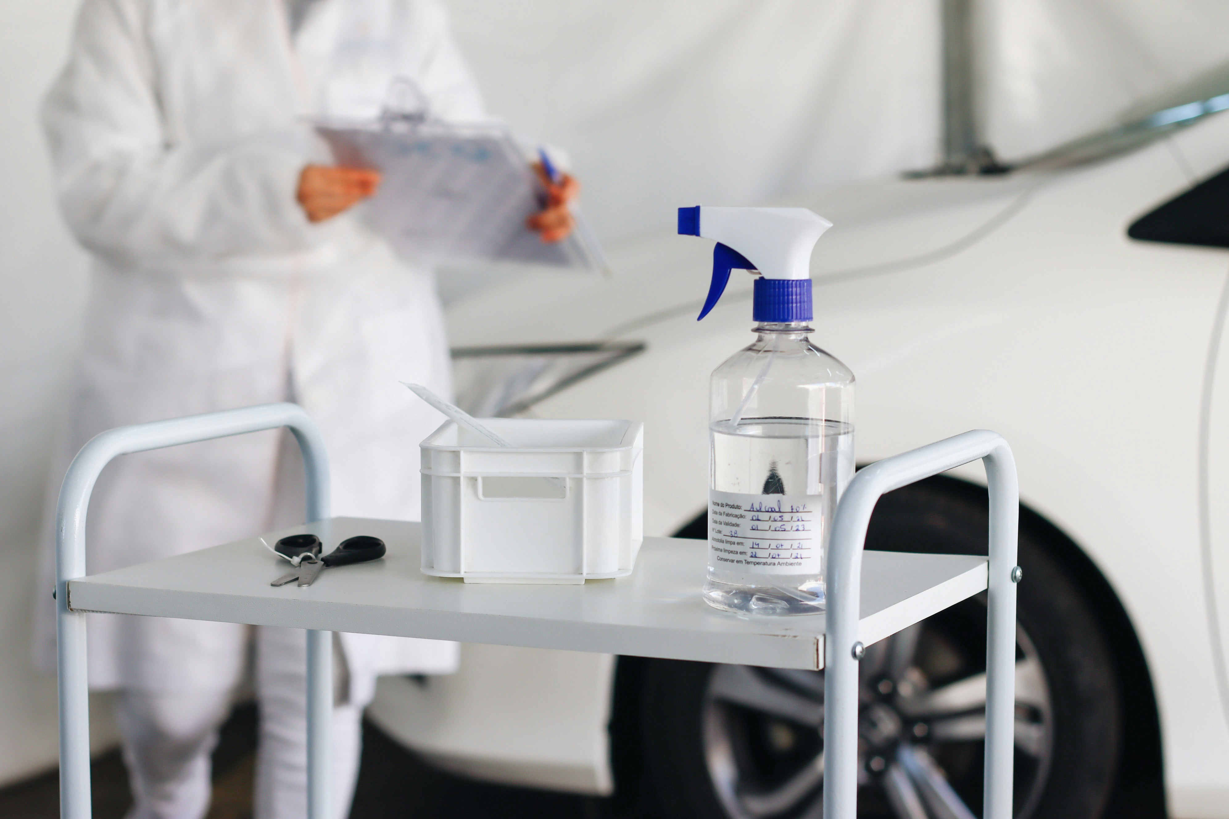 A disinfectant spray bottle and a white container on a mobile cart, with a person in a lab coat reviewing documents in the background. The setting suggests a focus on cleanliness and safety protocols.