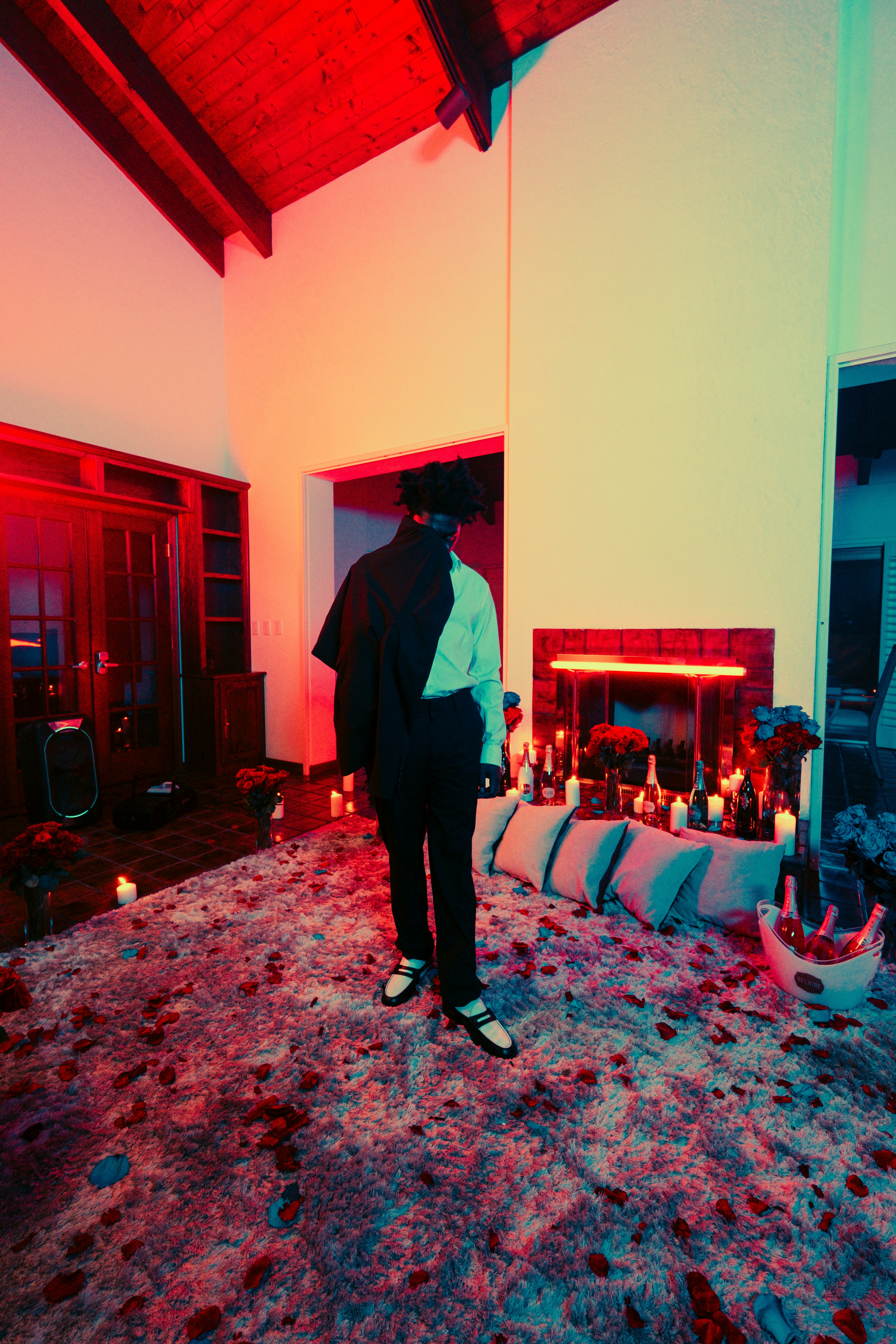 A figure stands in a dimly lit room adorned with candles and rose petals, evoking a sense of mystery and introspection.