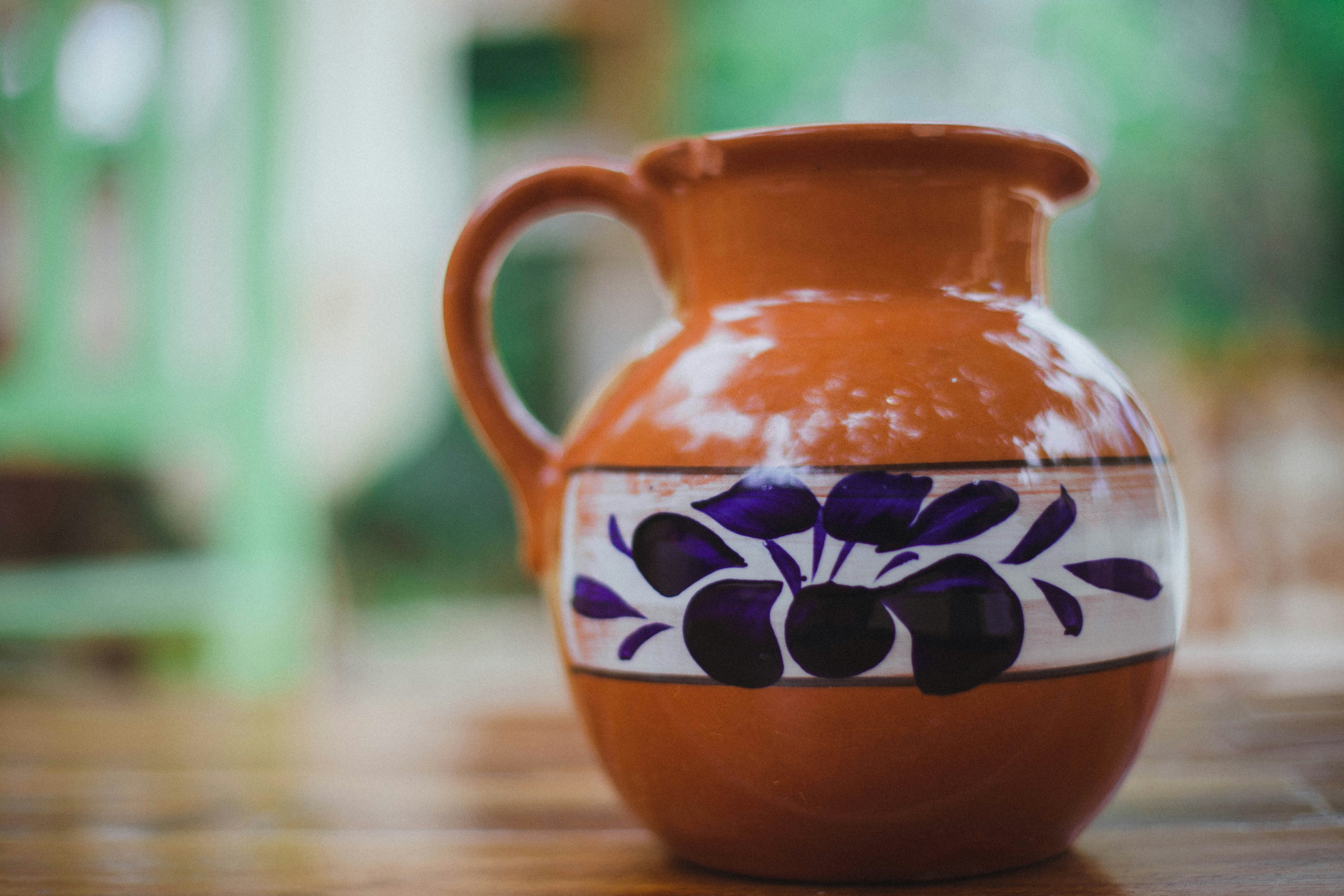 Handcrafted ceramic jug adorned with a delicate olive branch design, resting on a wooden surface. The background features a softly blurred green environment.