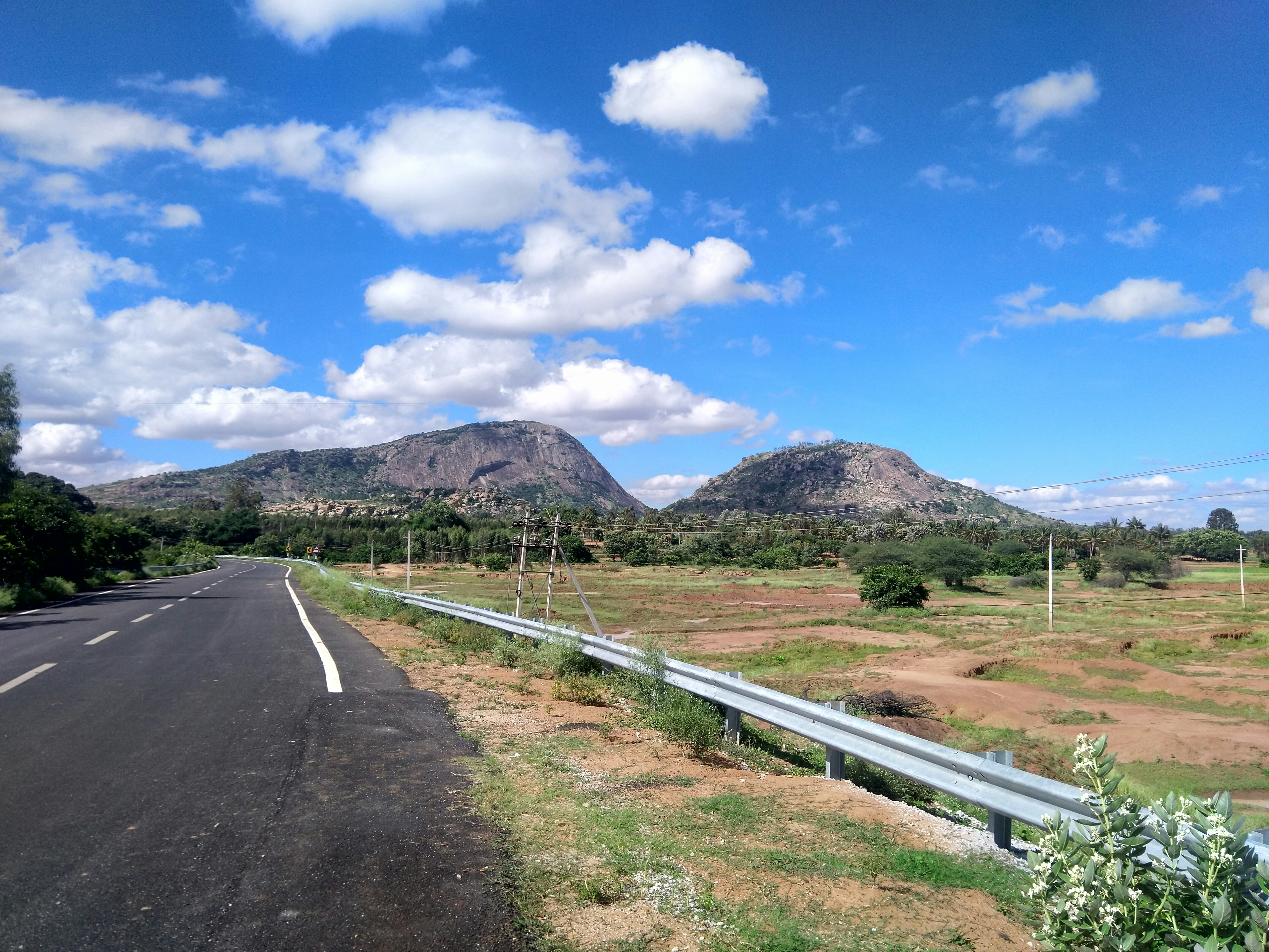 Photograph shows a rural highway curving toward distant hills beneath a bright, cloud-speckled sky. The road creates strong perspective and a sense of openness in the landscape.