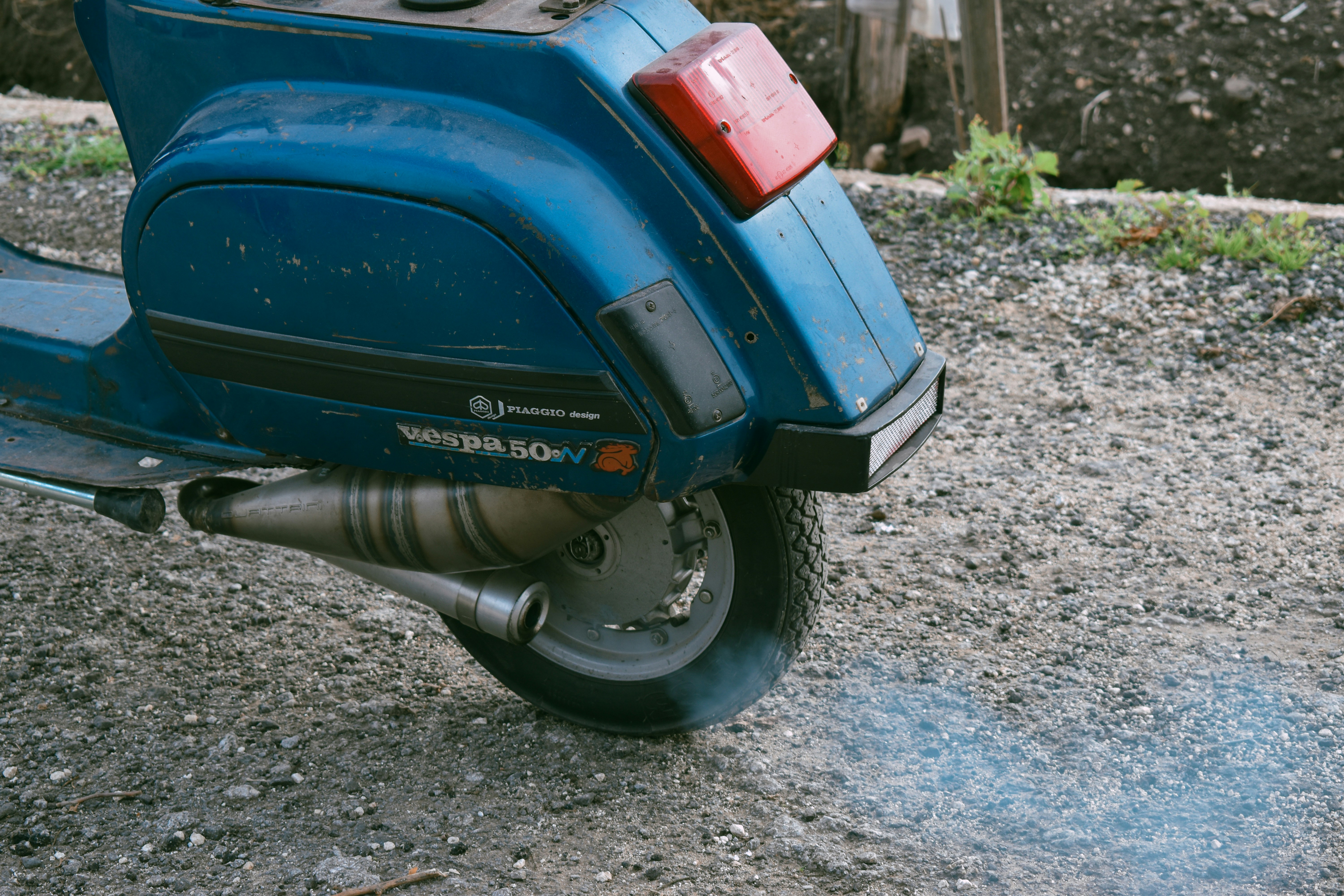 Azure Vespa Rear Quarter: Exhaust and Tail Light on Gravel