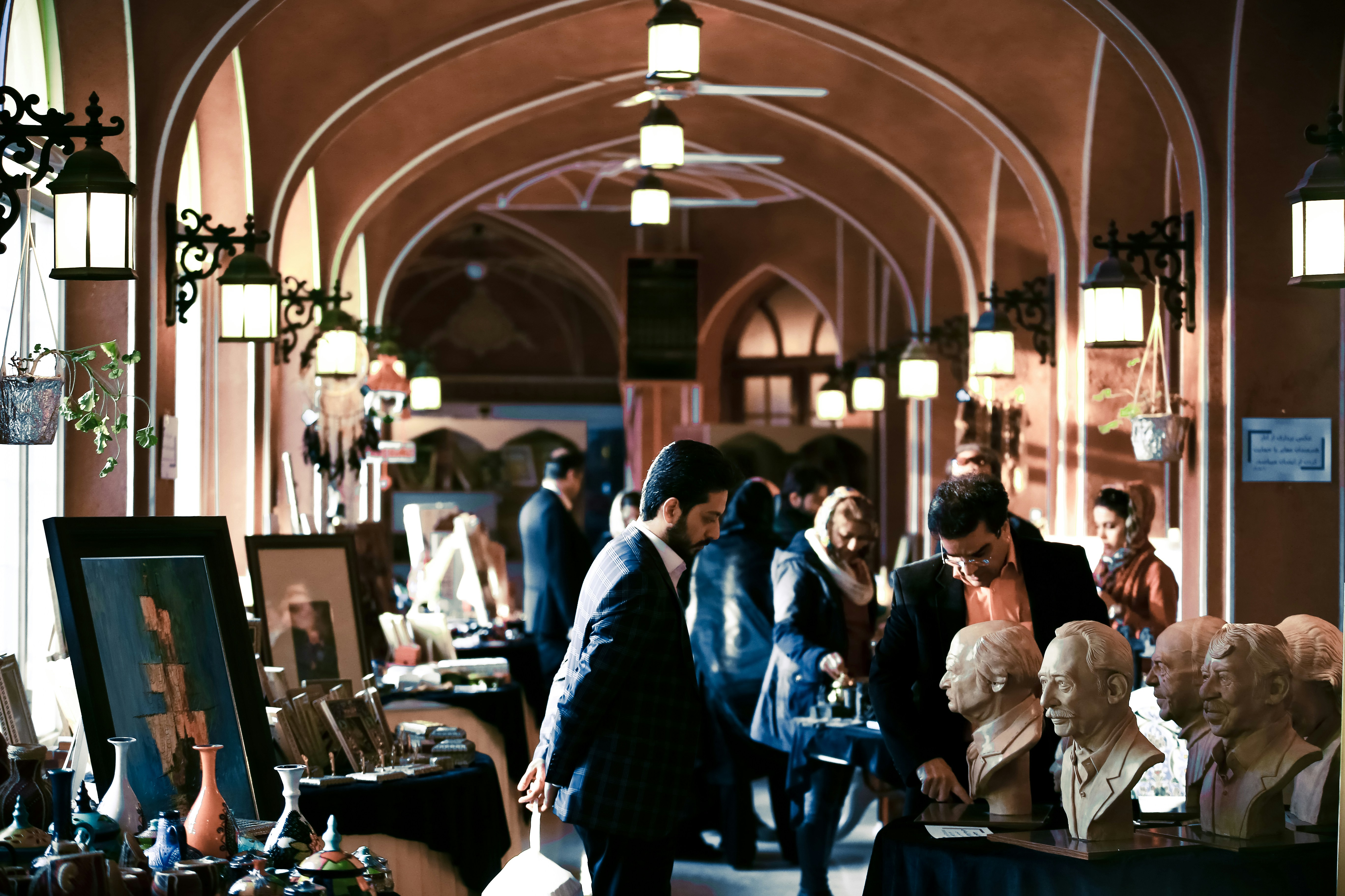 Artisans and visitors engage in a vibrant marketplace filled with sculptures and paintings under ornate arches. The warm ambiance enhances the artistic atmosphere.