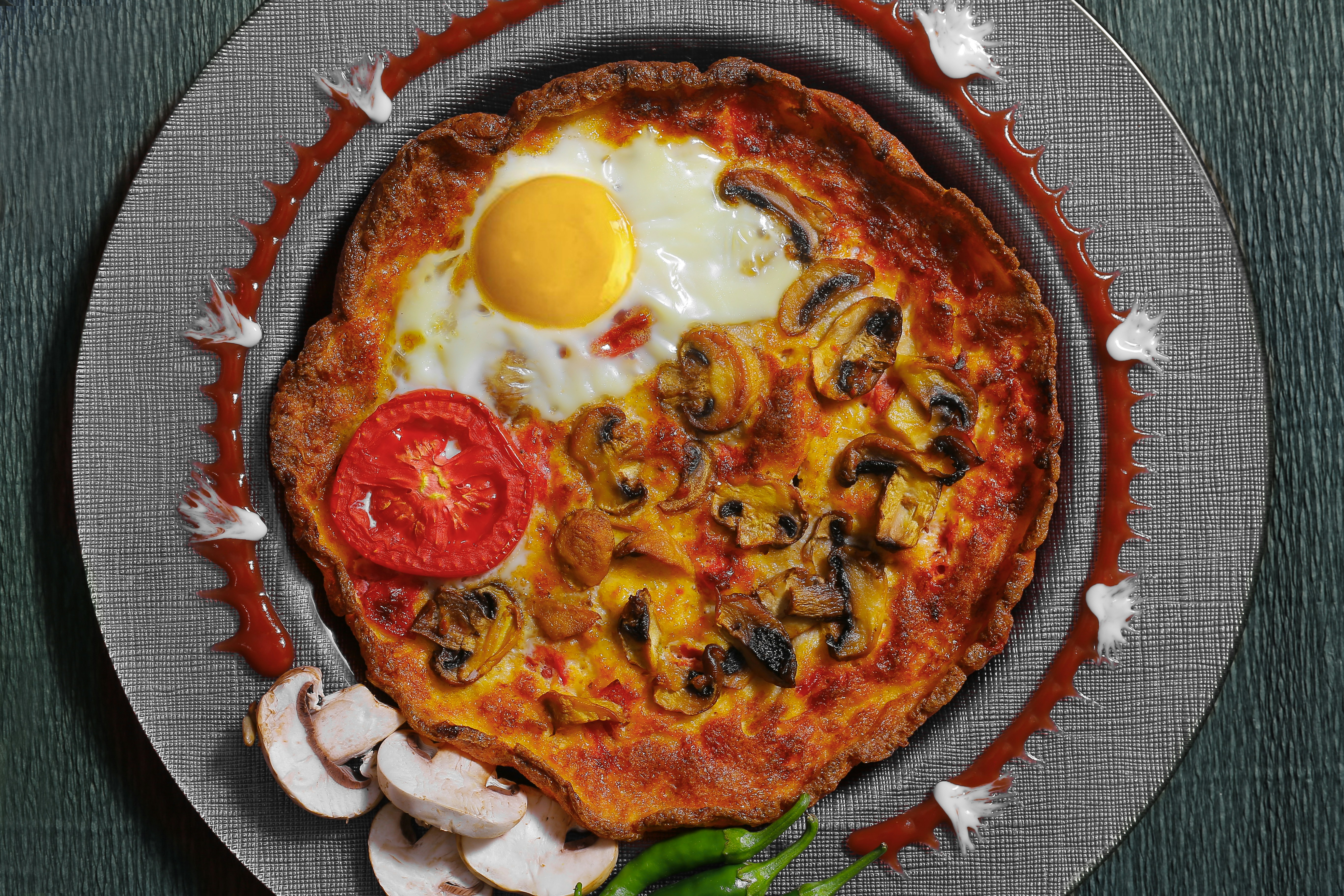 Breakfast pizza featuring a sunny-side-up egg, mushrooms, and fresh vegetables, elegantly arranged on a textured plate.