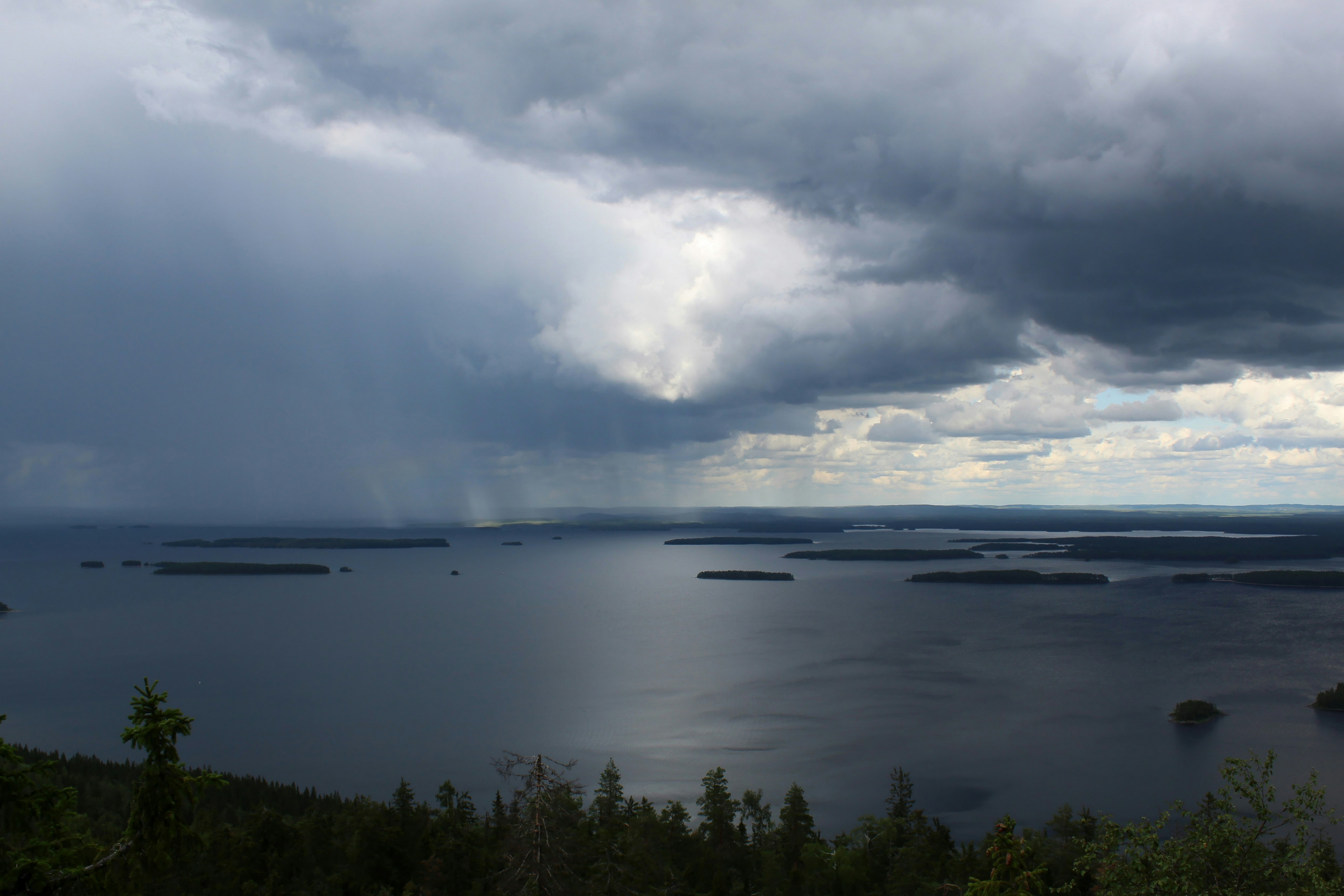 Dark clouds loom over a serene lake dotted with islands, hinting at an approaching storm. The contrast between the water's calm surface and the turbulent sky creates a dramatic atmosphere.