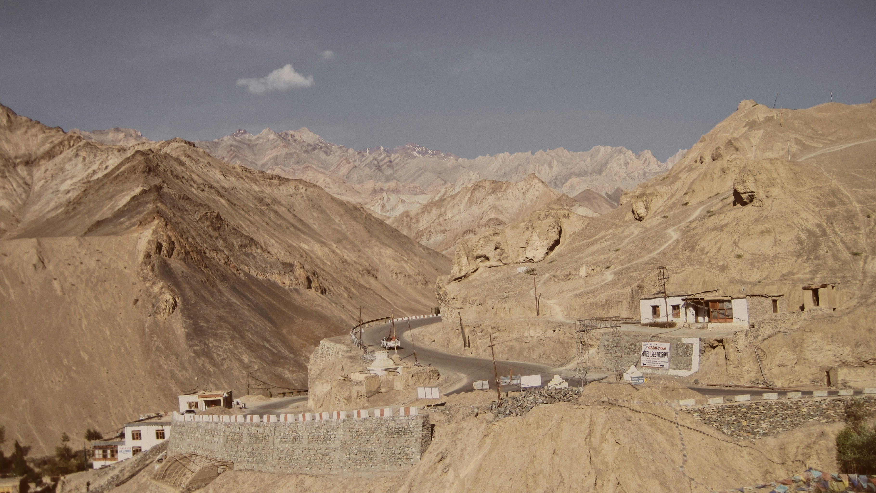 A desert valley scene features a sinuous road winding between wind-sculpted hills and distant mountains. Small white buildings line the hillside, providing human scale in the stark landscape.