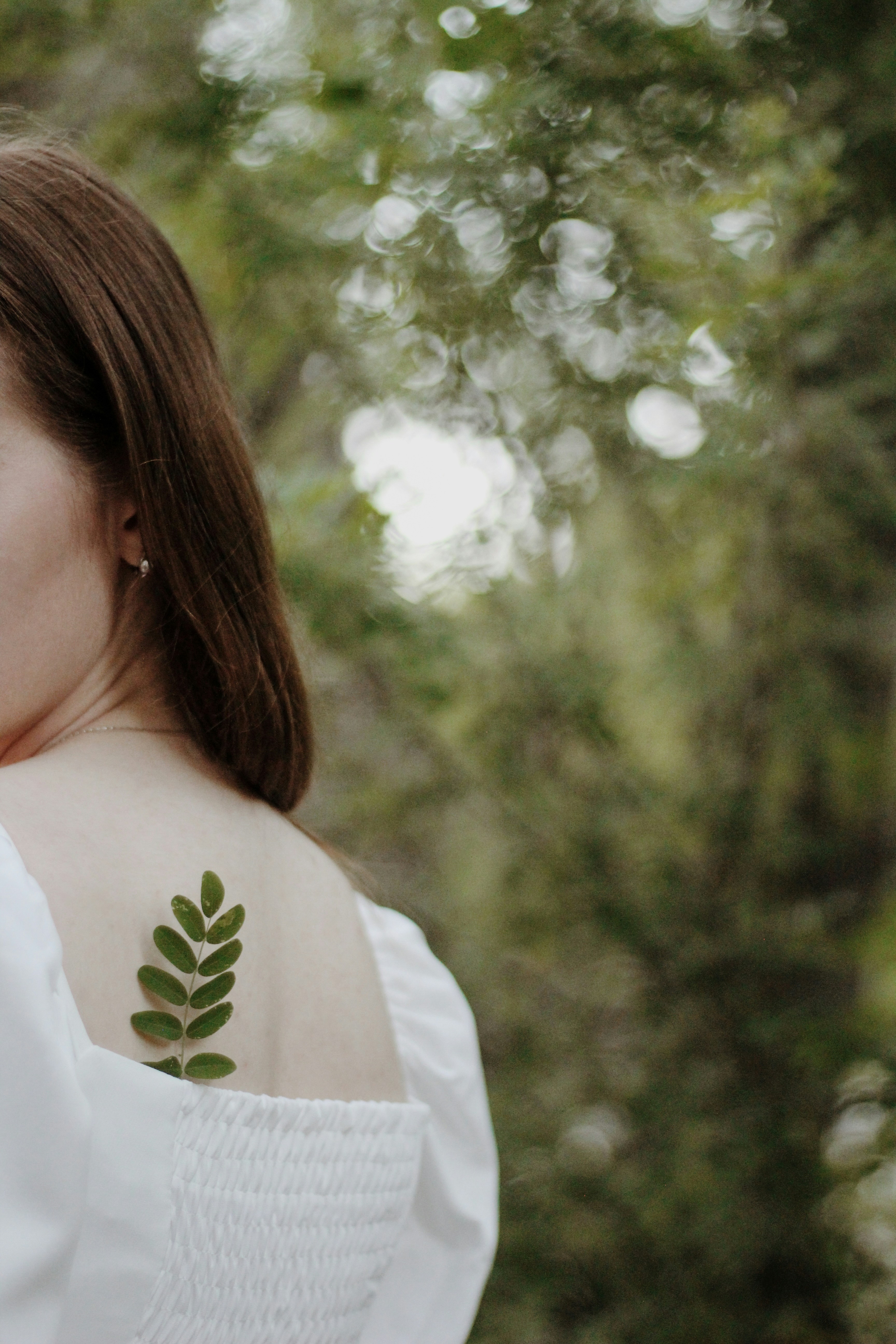 A woman gazes over her shoulder, revealing a delicate leaf tattoo against a blurred natural backdrop. The soft focus enhances the connection between the subject and nature.
