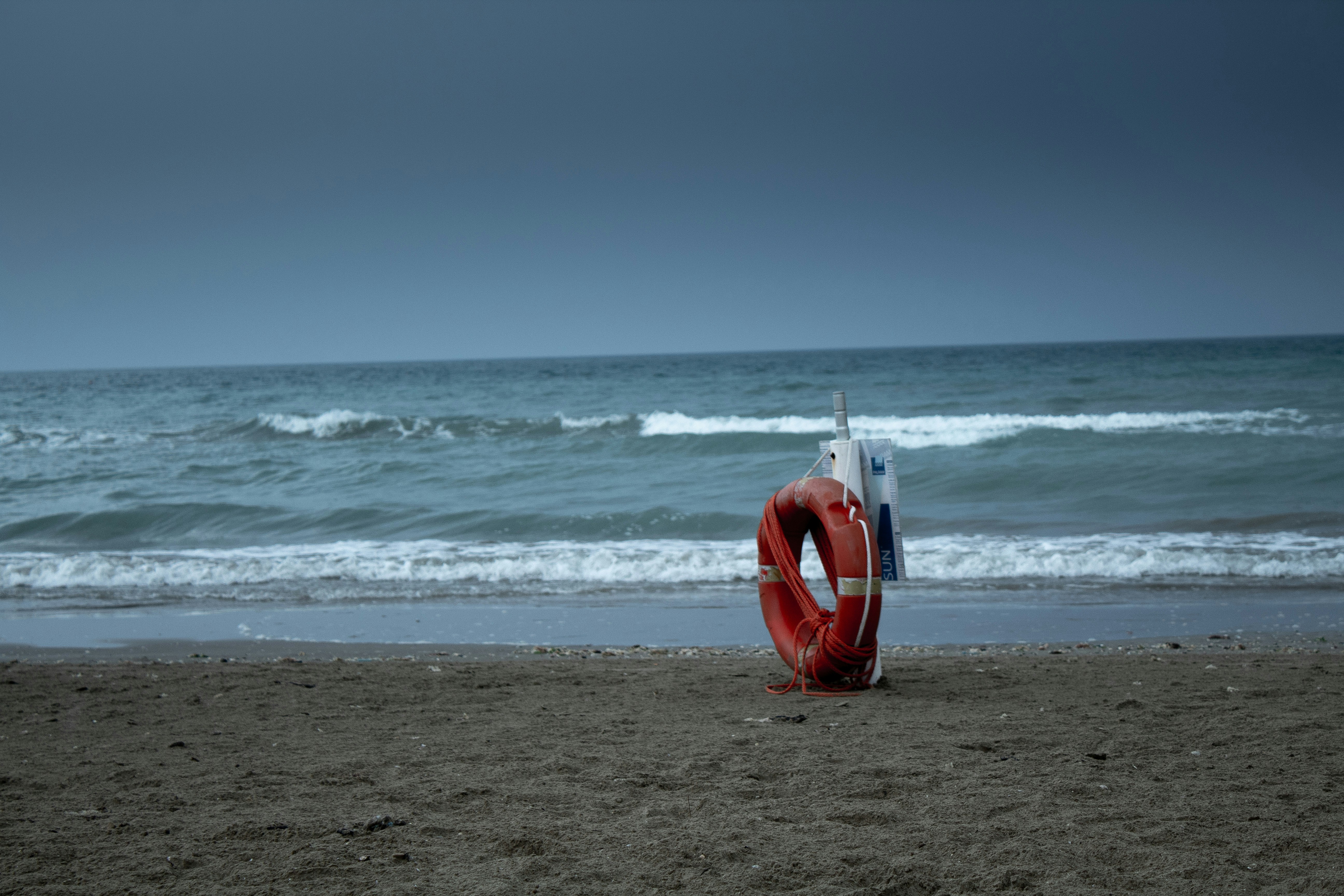 Lifebuoy positioned on sandy beach beside calm waves under a moody sky.