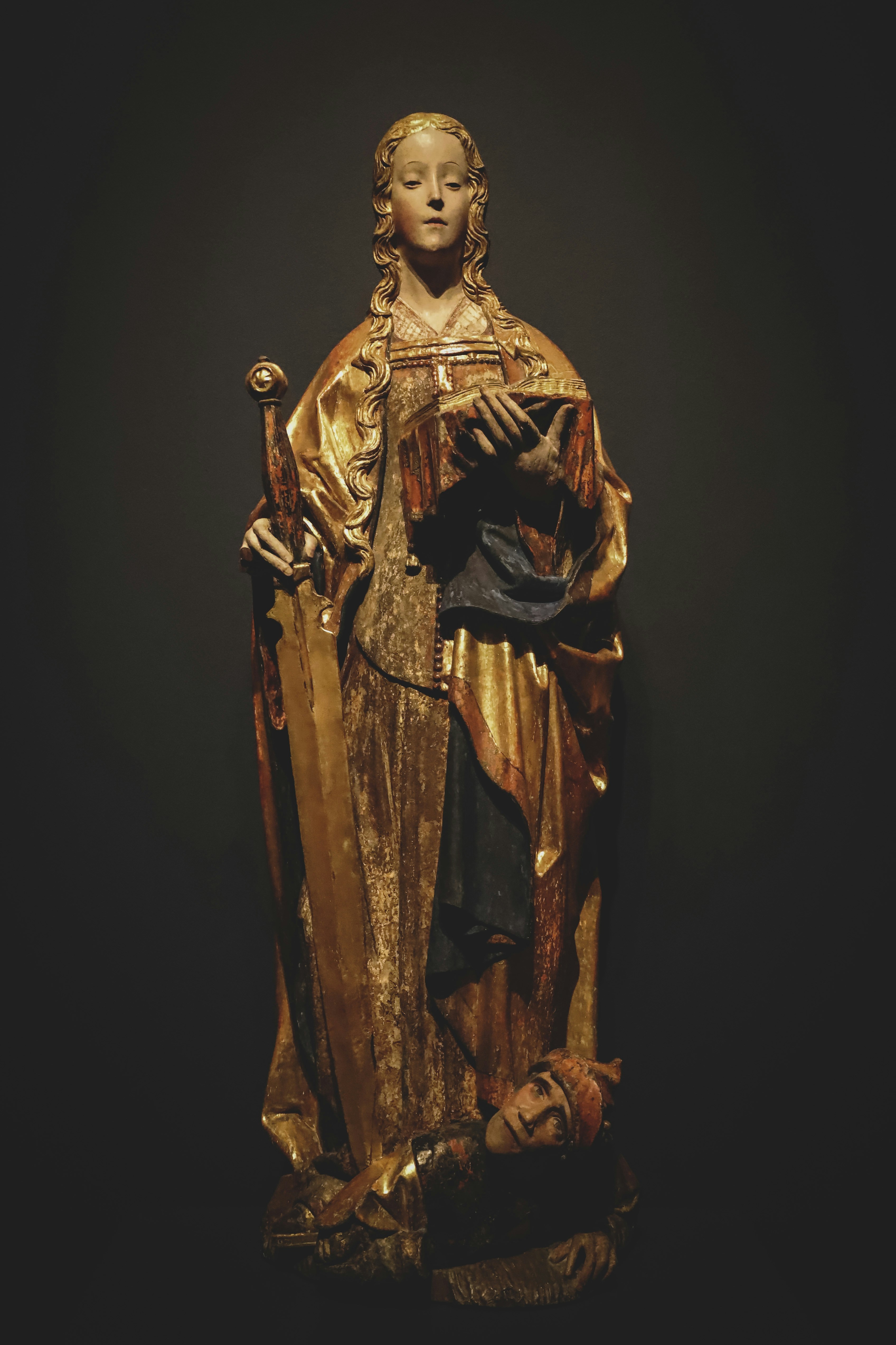 Intricately carved wooden statue of a saint holding a sword and book, with a defeated figure at her feet. The rich textures and colors evoke a sense of historical reverence.