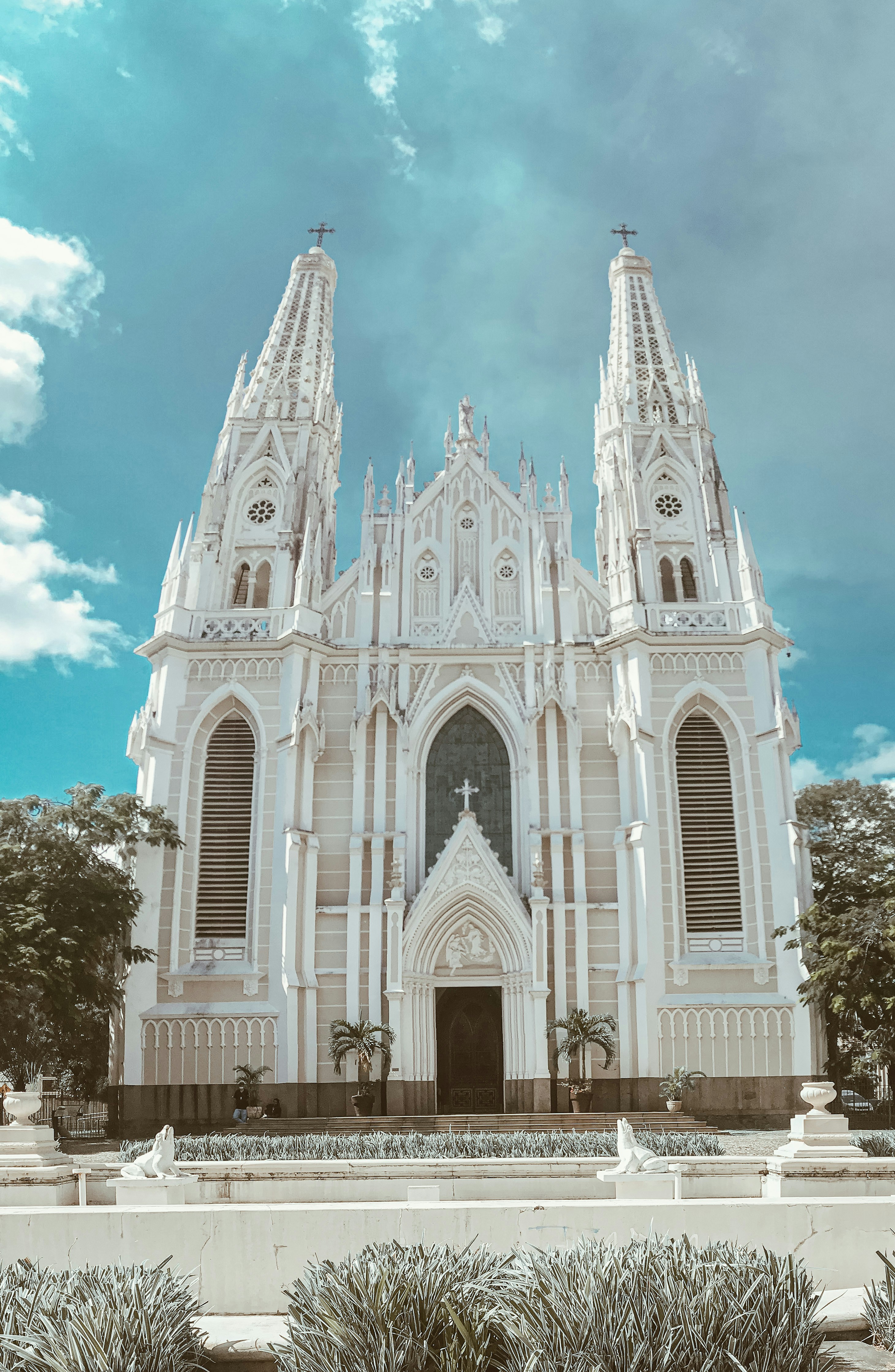 Gothic-style church with towering spires and intricate details under a vibrant blue sky. Lush greenery frames the scene.