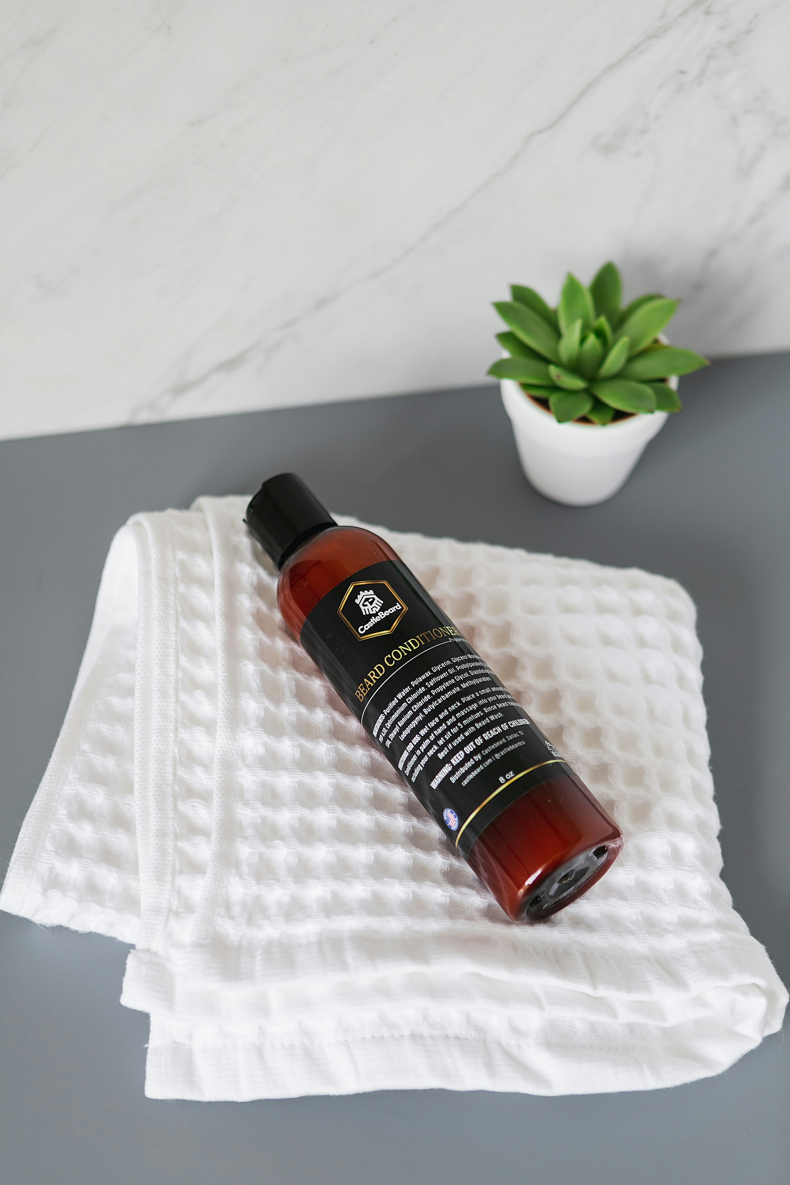 Beard conditioner bottle resting on a textured white towel beside a small potted plant. The minimalist setup emphasizes grooming and self-care.