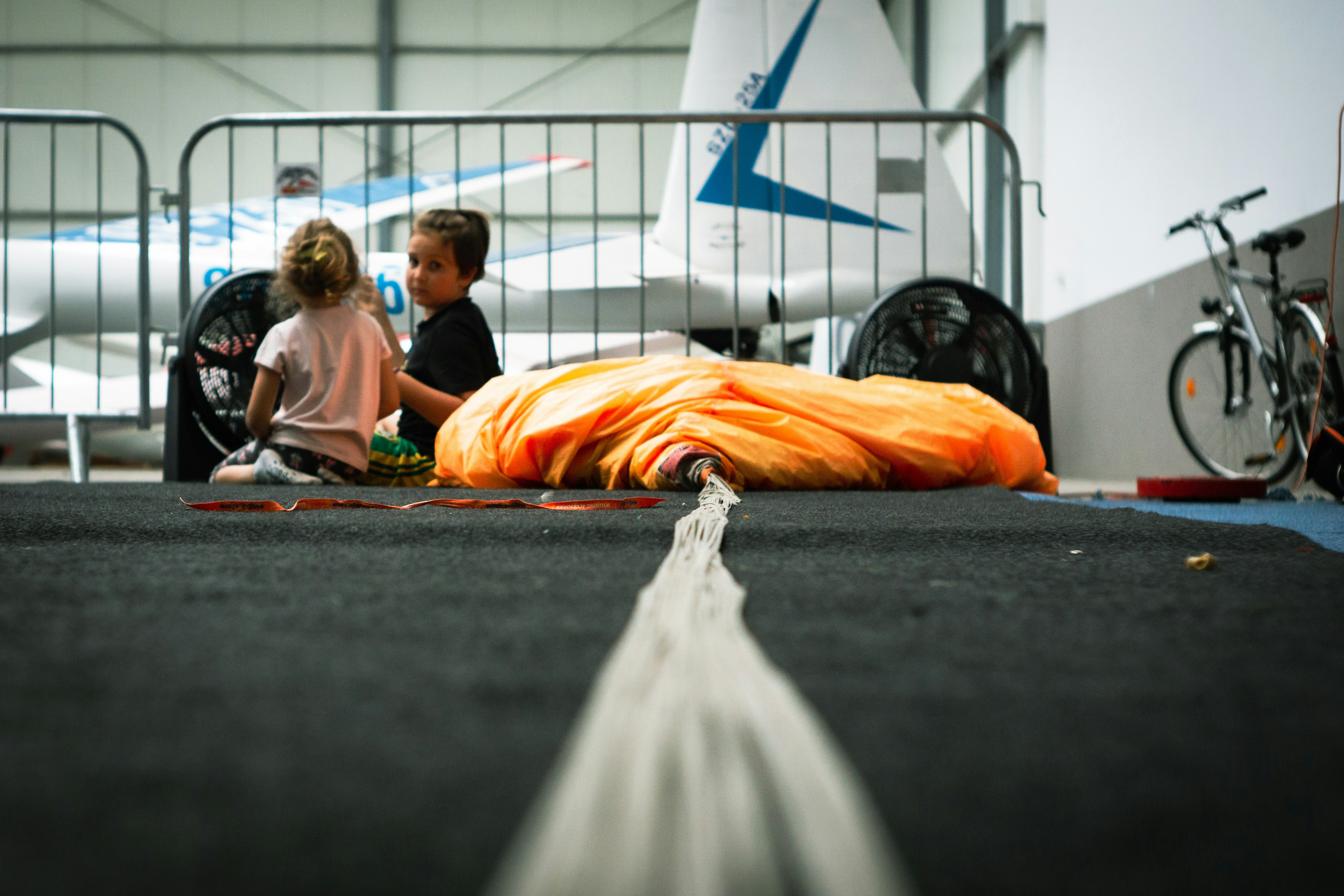 Hidden in the Hangar: Two children beside a bright orange bag in an aircraft workshop