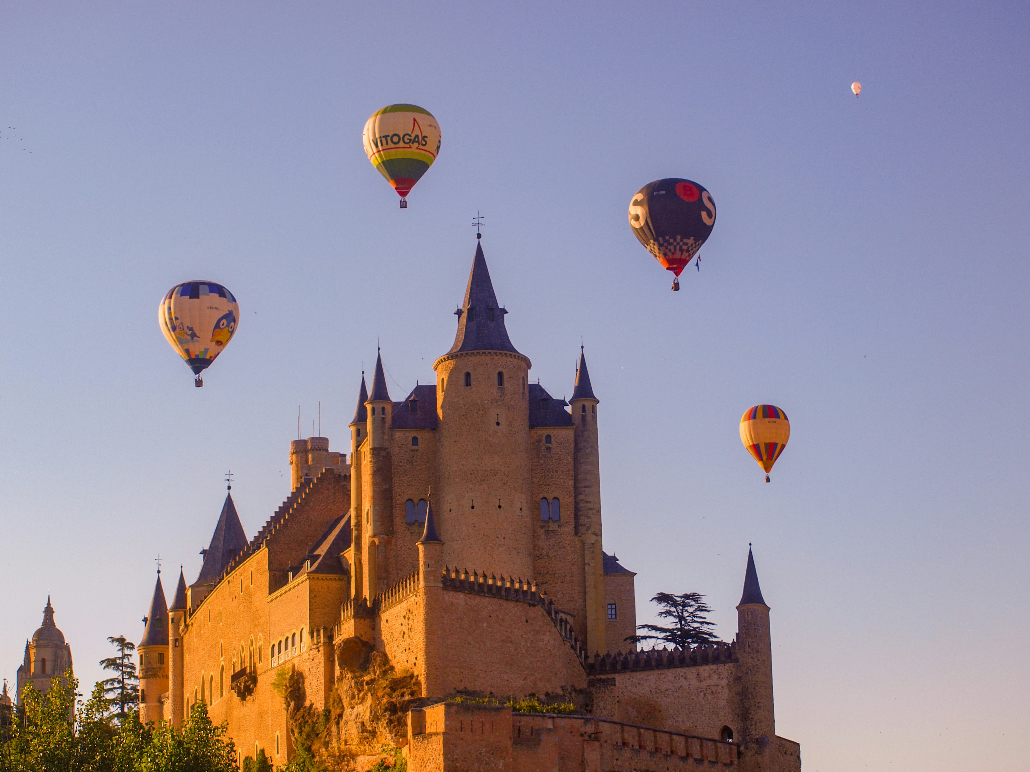 Colorful hot air balloons soar over a historic castle under a clear blue sky.