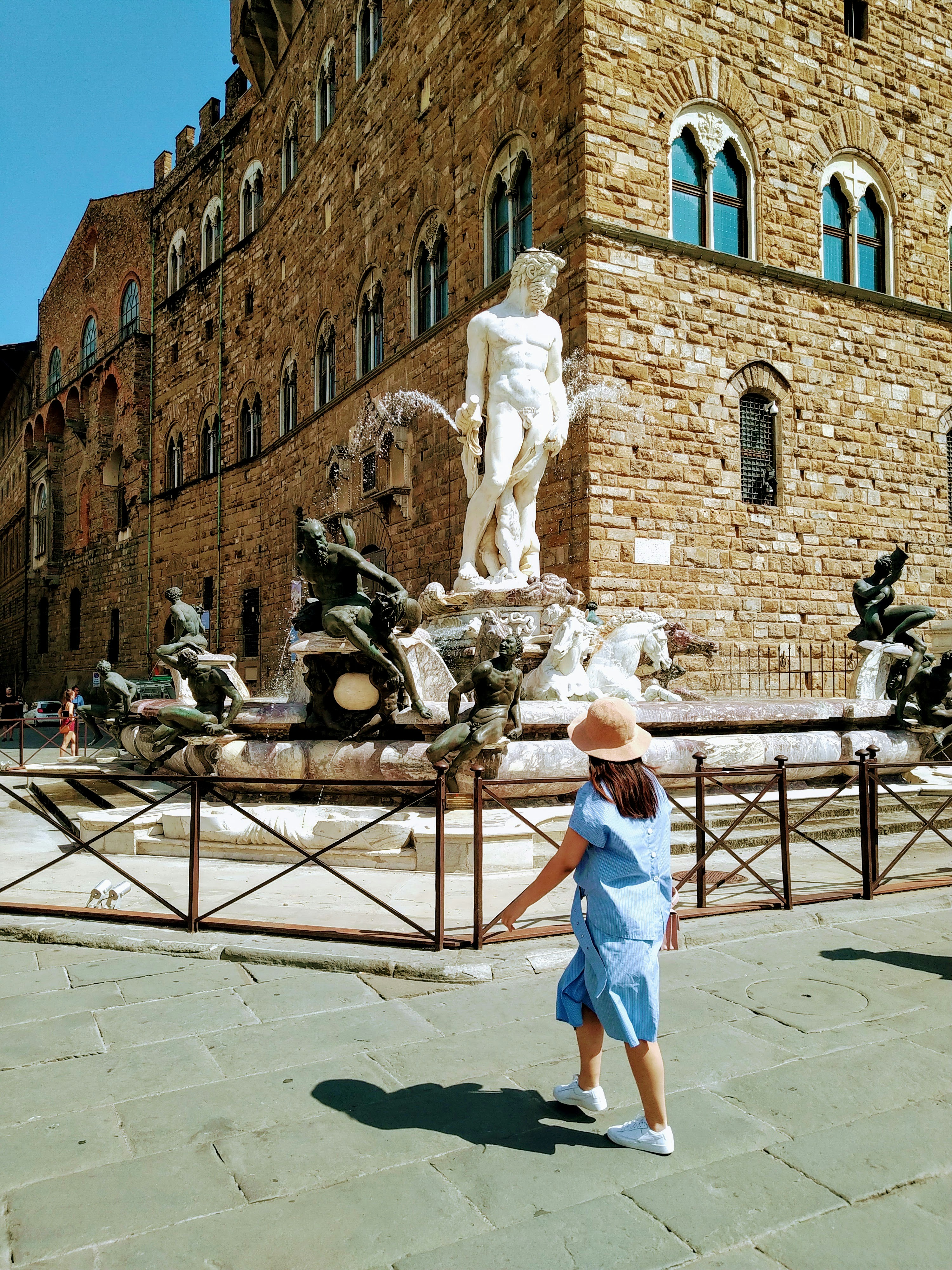 A woman in a blue dress and sunhat walks past a classical fountain with a central marble statue and bronze figures, set against a sunlit brick building.
