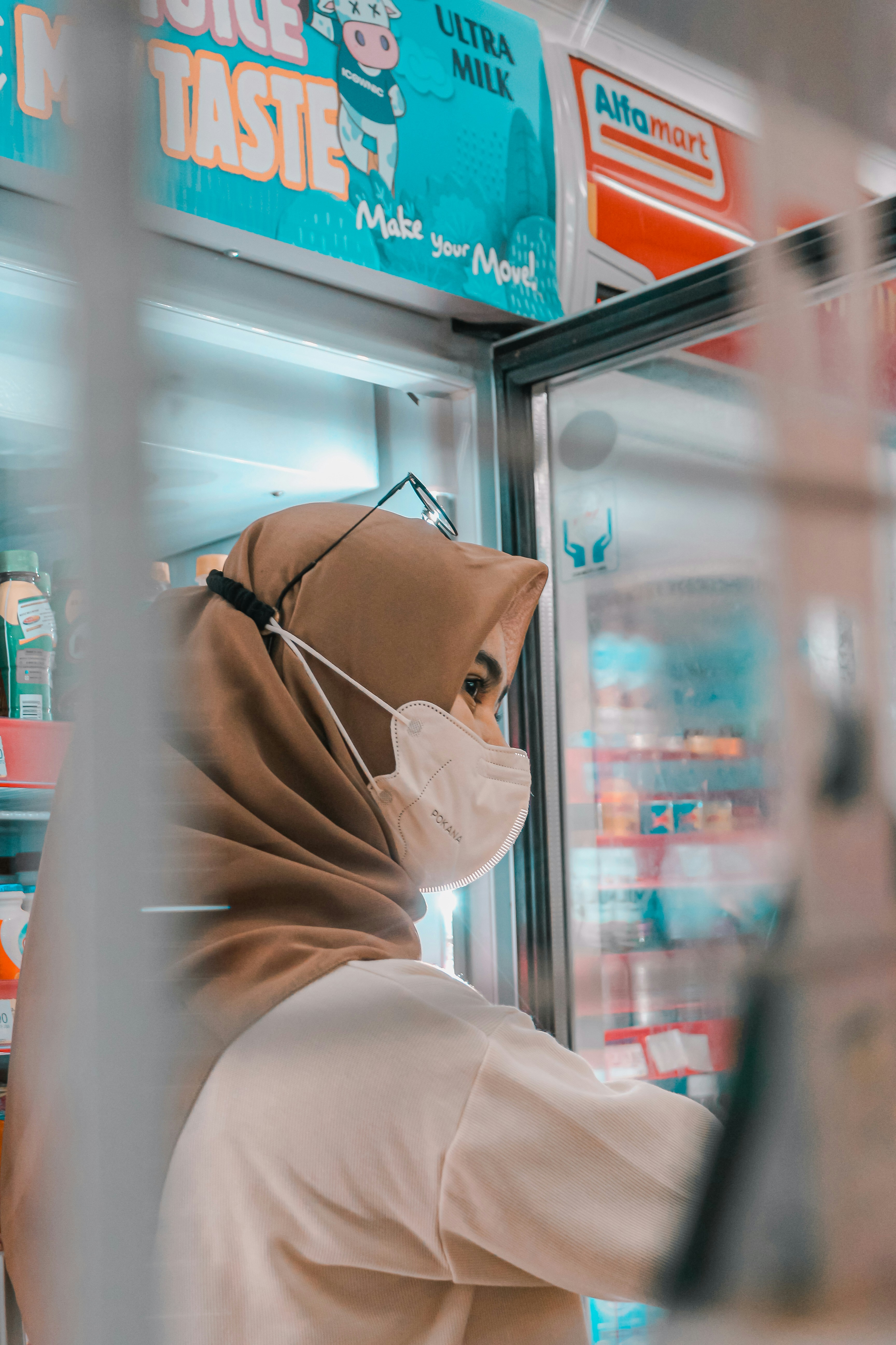 Woman in a beige hijab selecting items from a refrigerator in a convenience store, framed by glass and shelves. 
