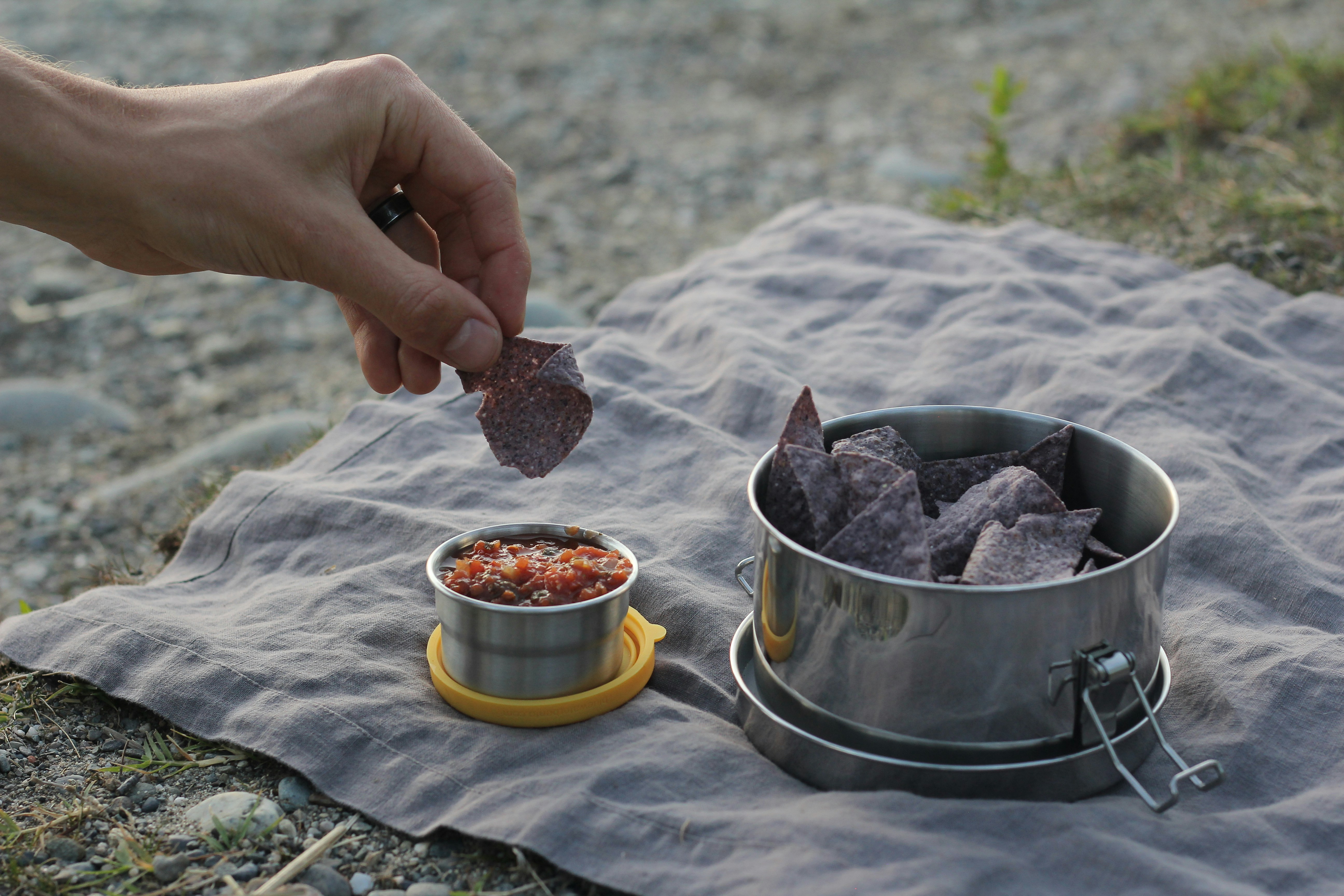 Hand reaching for a blue corn tortilla chip above a small bowl of salsa, set on a textured cloth amidst a natural outdoor setting.