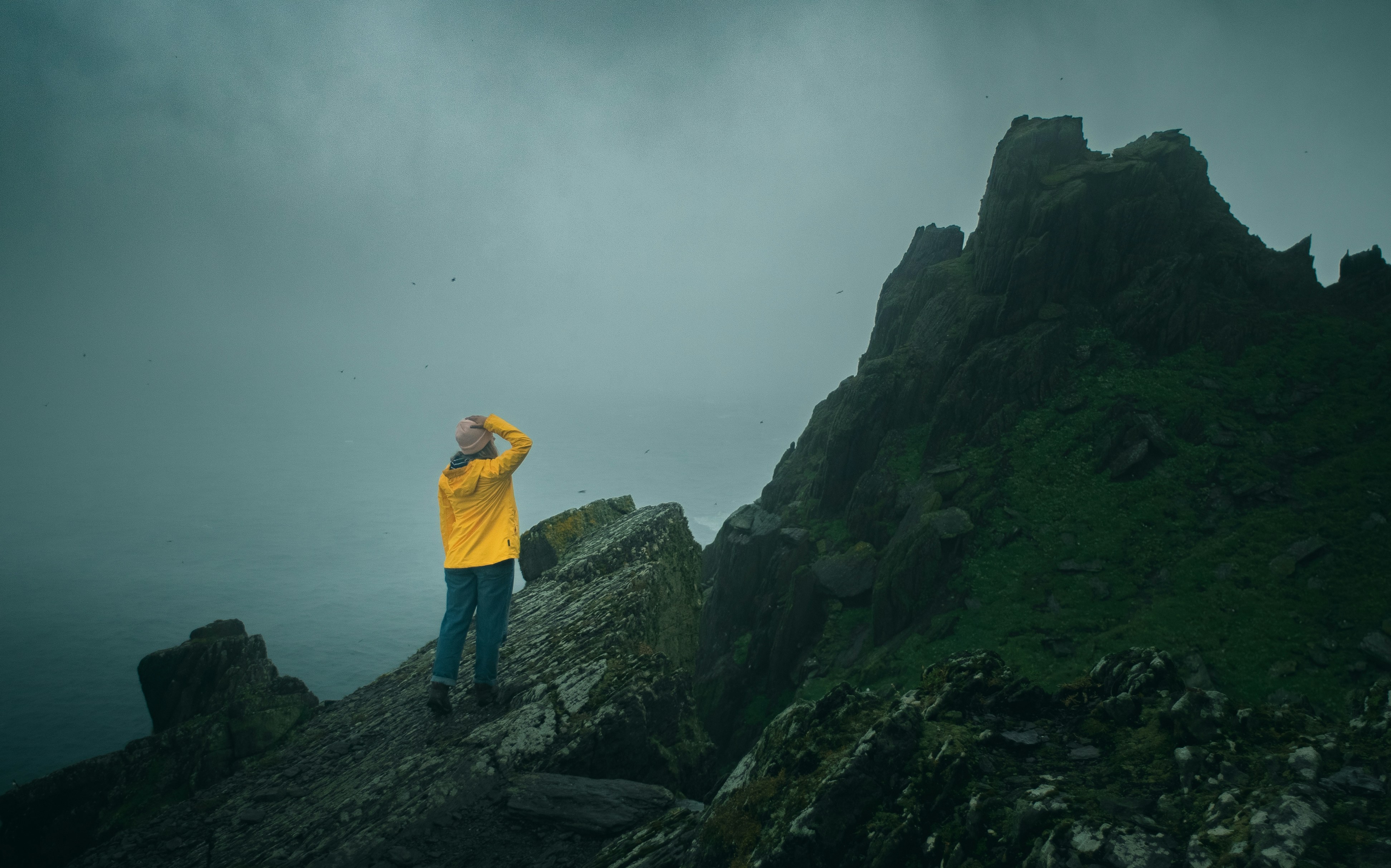 Person in a yellow jacket on rocky cliff with misty backdrop.