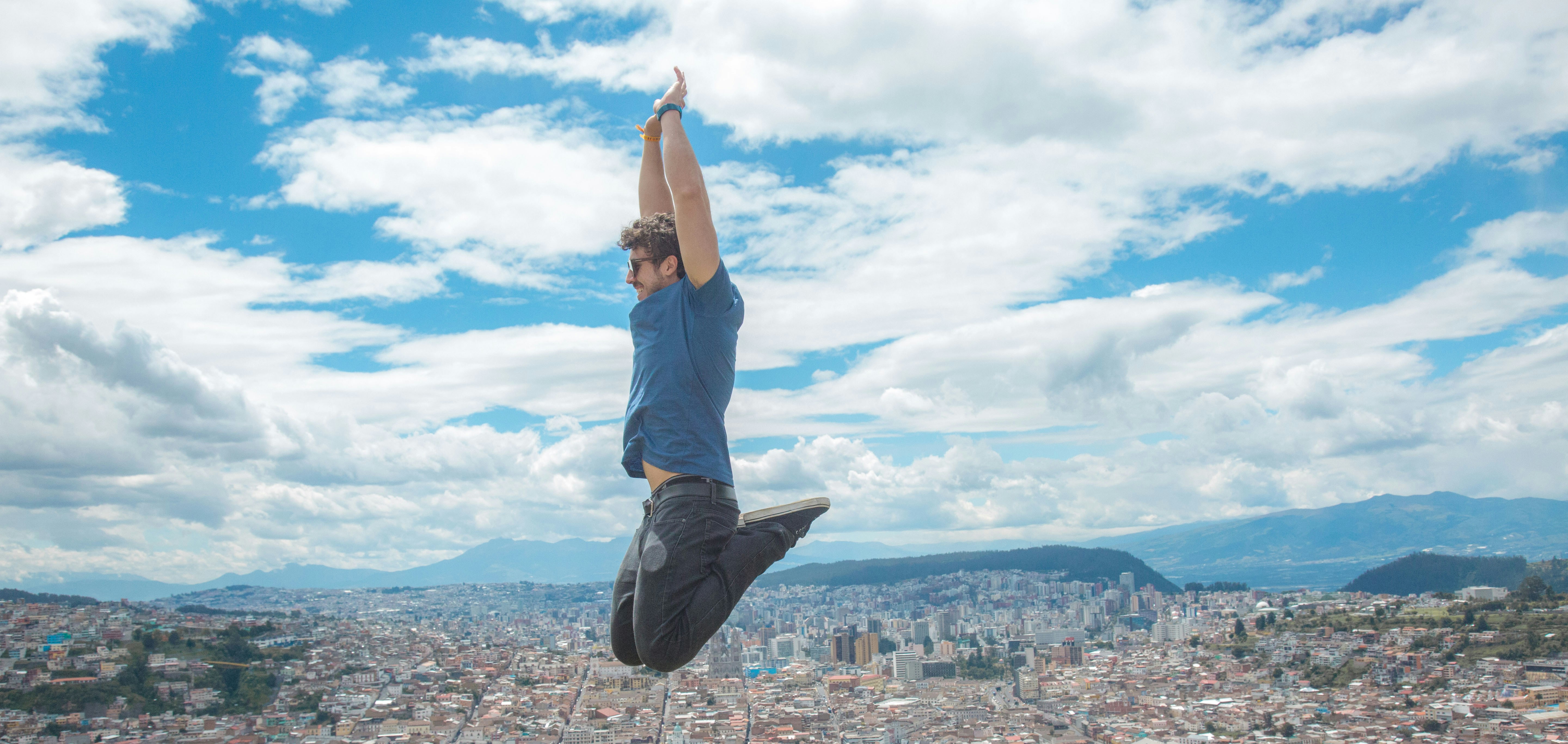 Person in blue tank top and jeans jumping with arms raised against a city skyline and cloudy sky.