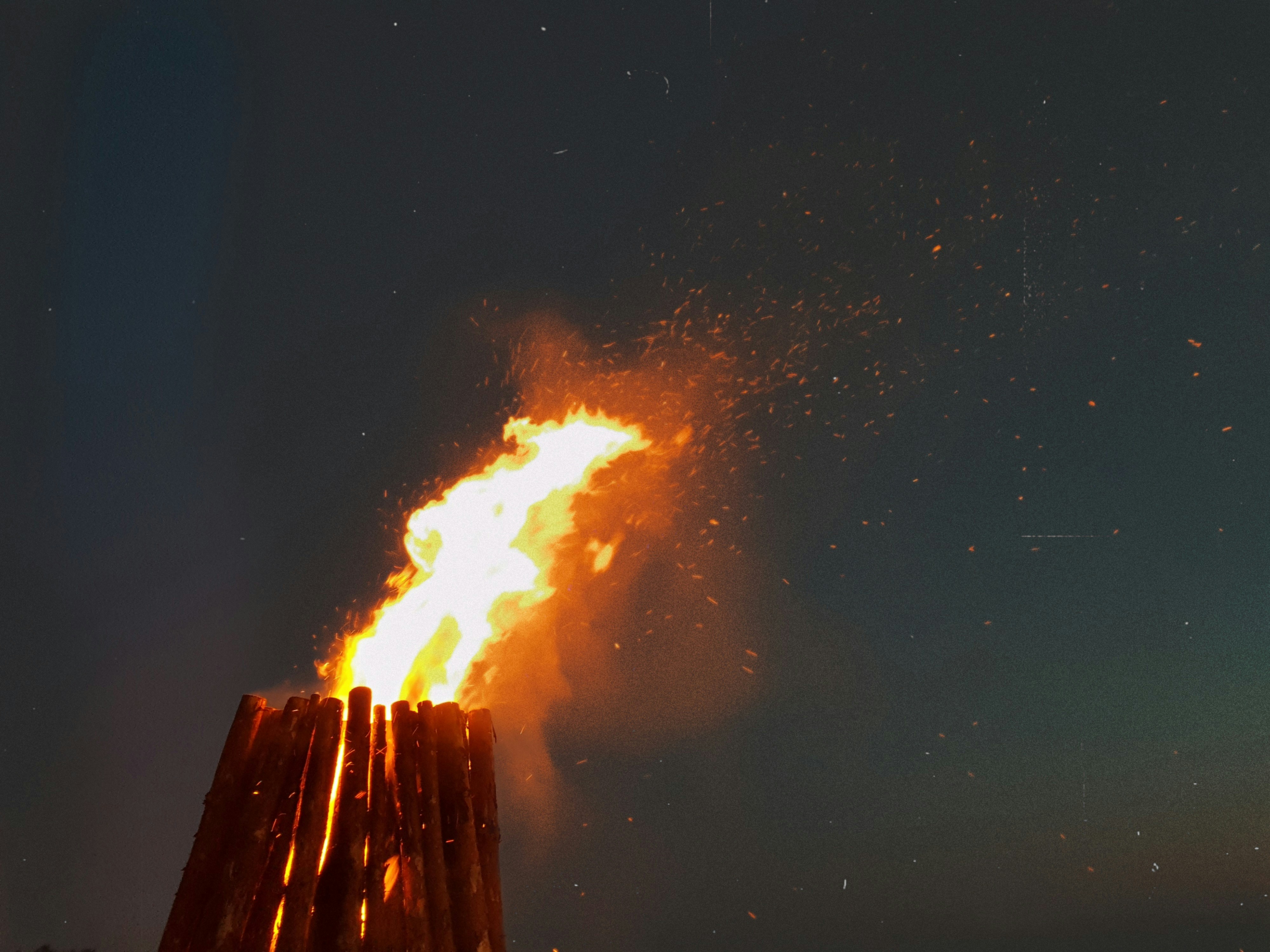 A towering bonfire erupts with vibrant flames and flying embers against a twilight sky.