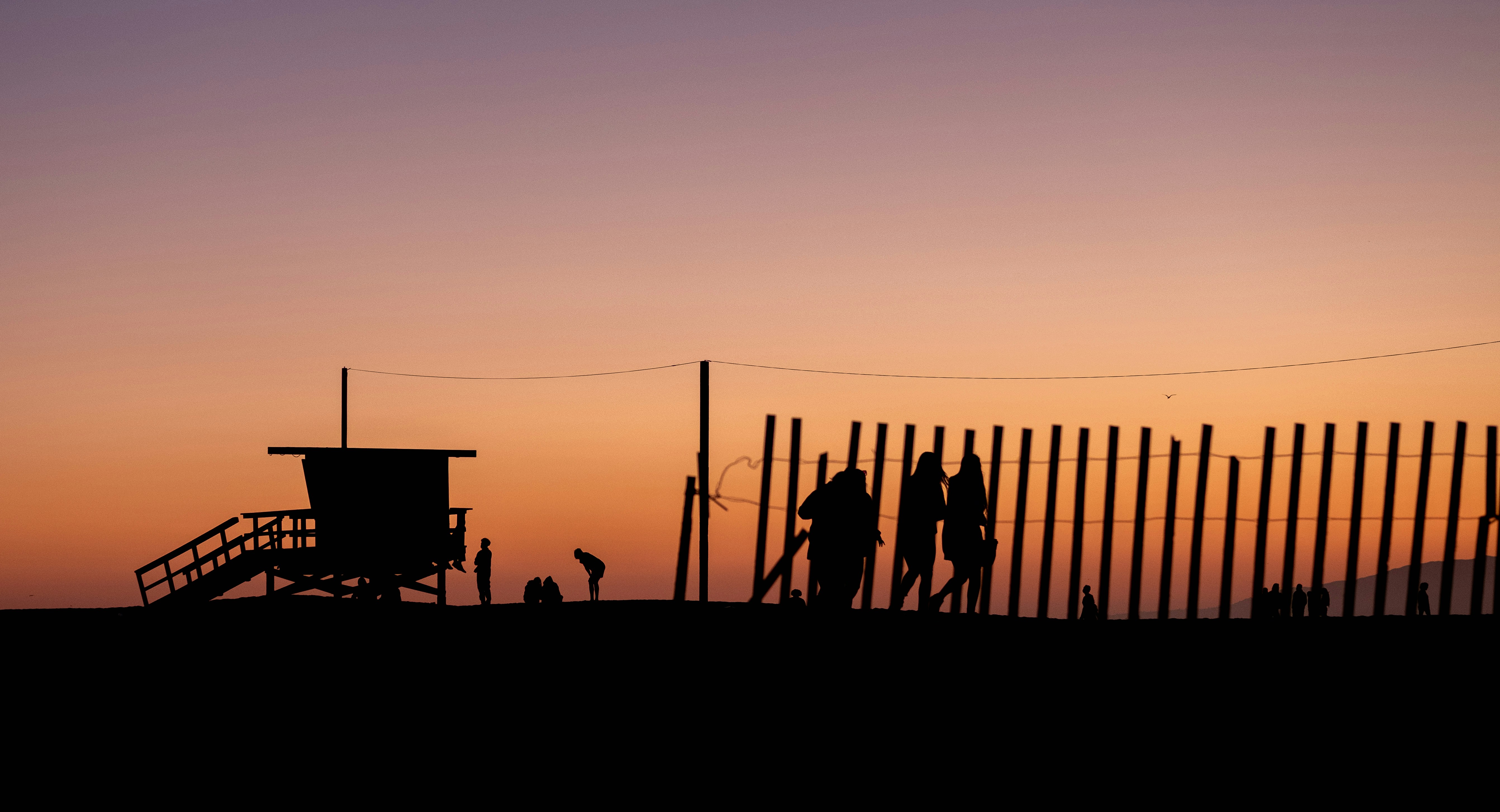 Lifeguard tower and silhouettes of people walking along a beach at sunset, framed by vertical wooden poles. 