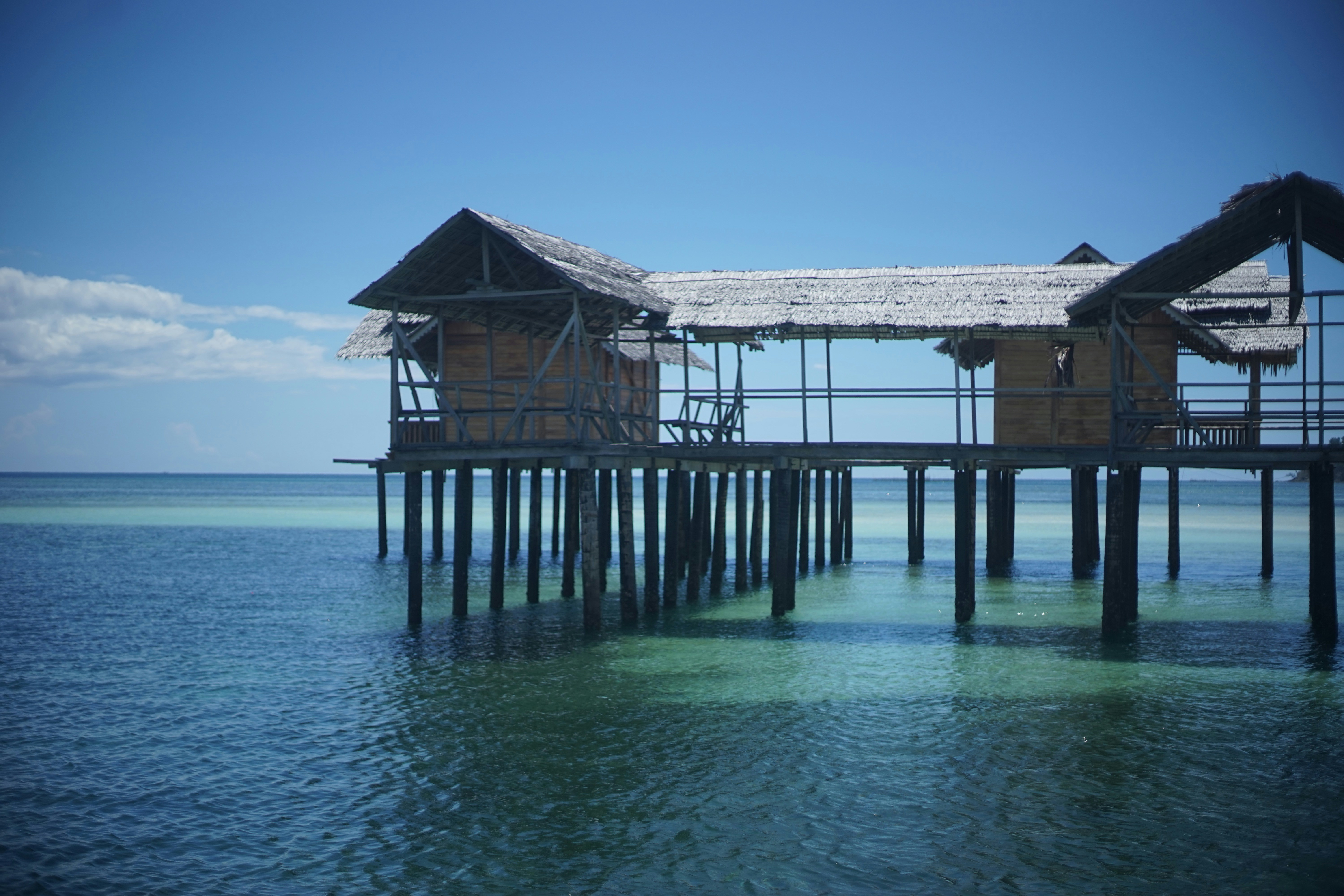 Wooden stilt house extending over clear turquoise sea under a bright blue sky.