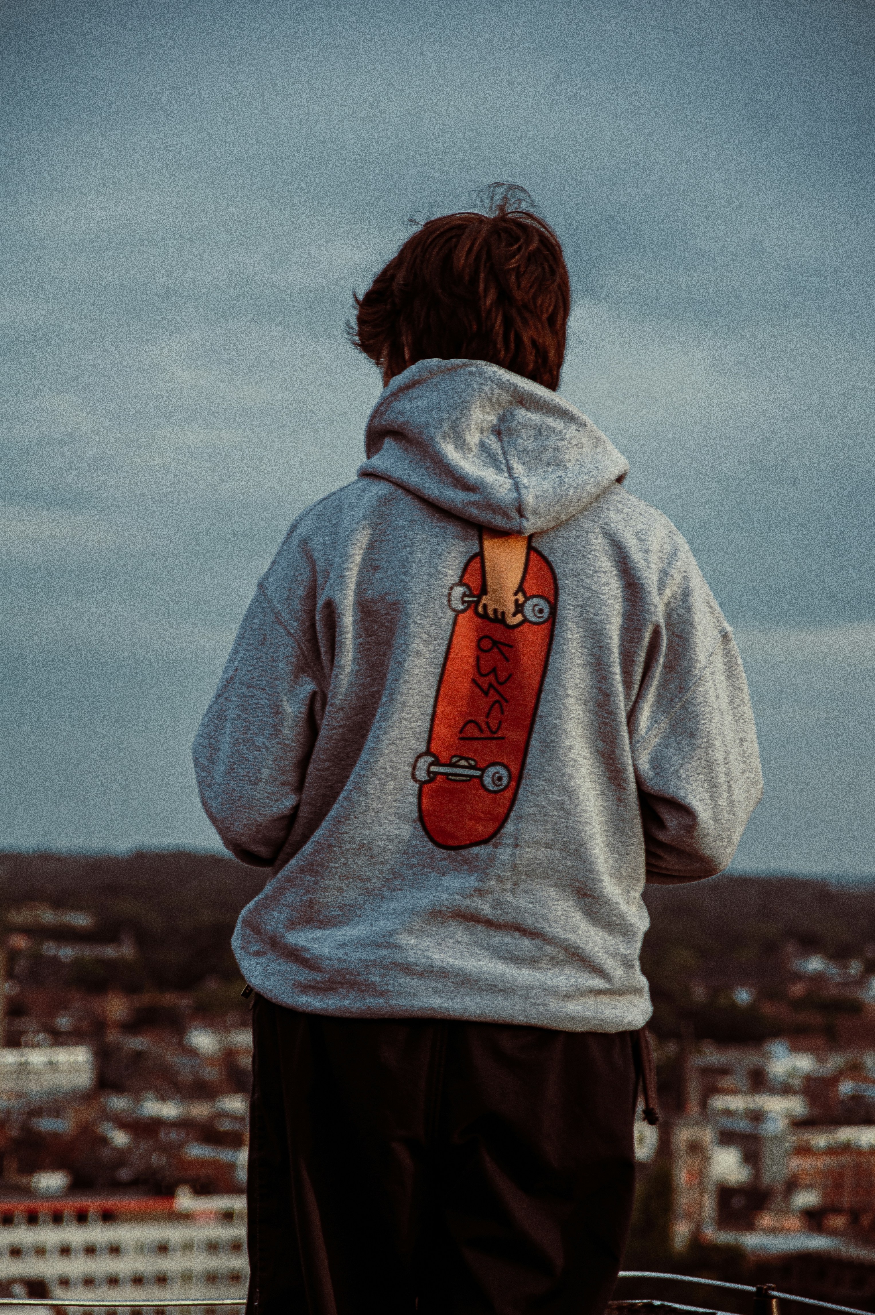 Individual wearing a hoodie with a skateboard design stands overlooking a cityscape, embodying the spirit of urban exploration.
