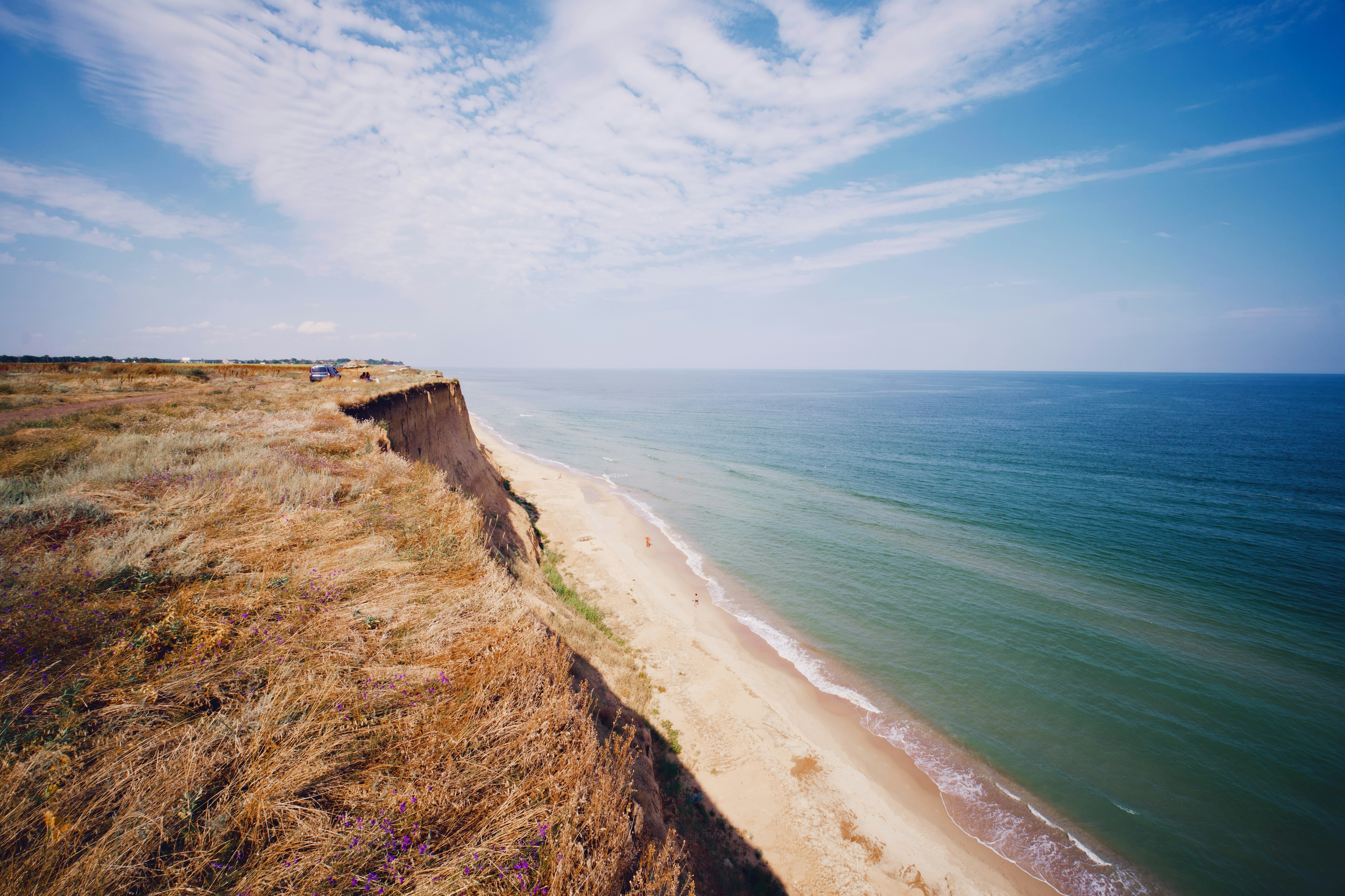 Dramatic coastal cliff overlooking a sandy beach and expansive sea under a partly cloudy sky.