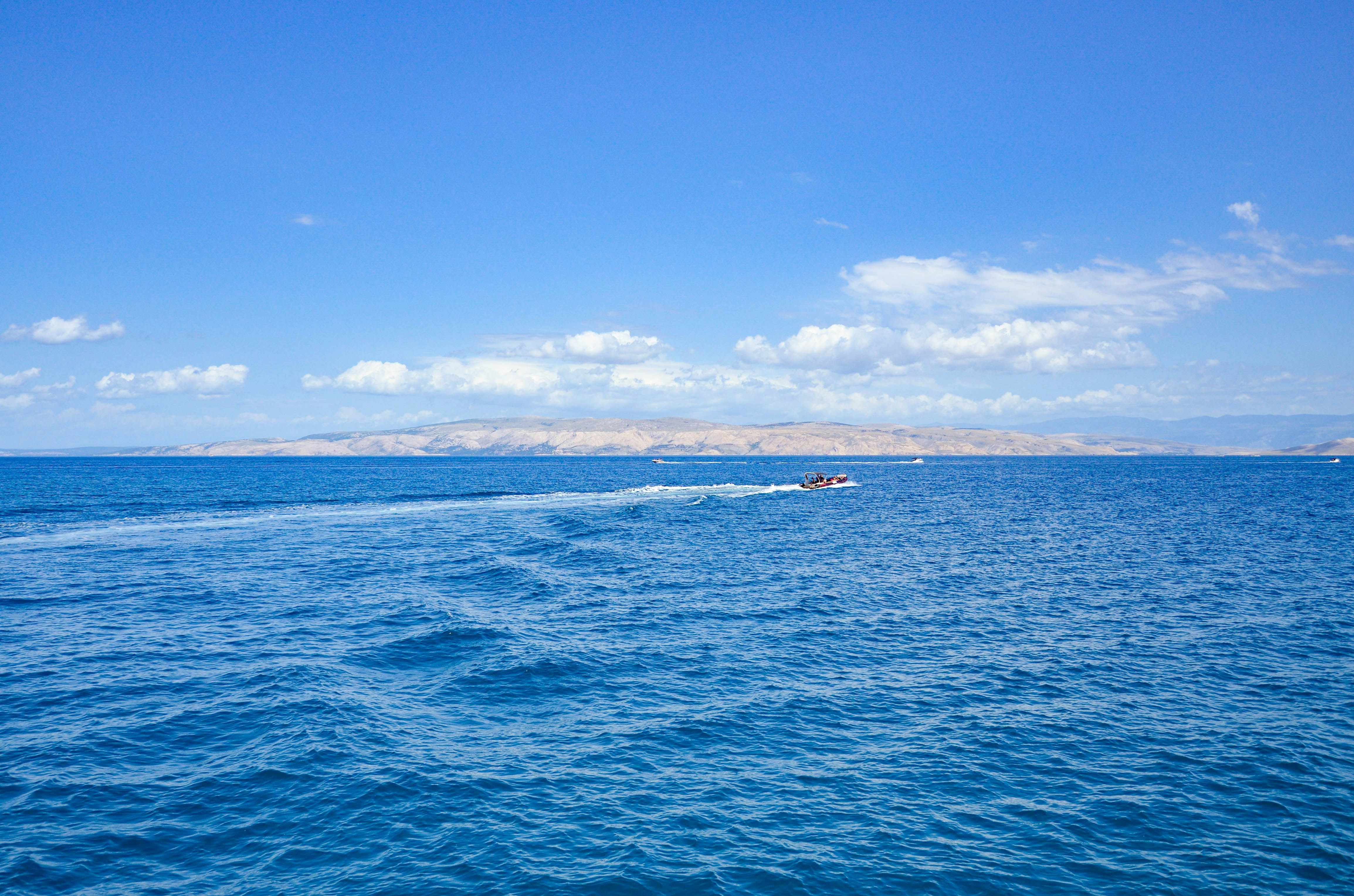 A speedboat glides across the calm sea, leaving gentle ripples in its wake under a clear blue sky. The distant landmass adds depth to the tranquil seascape.