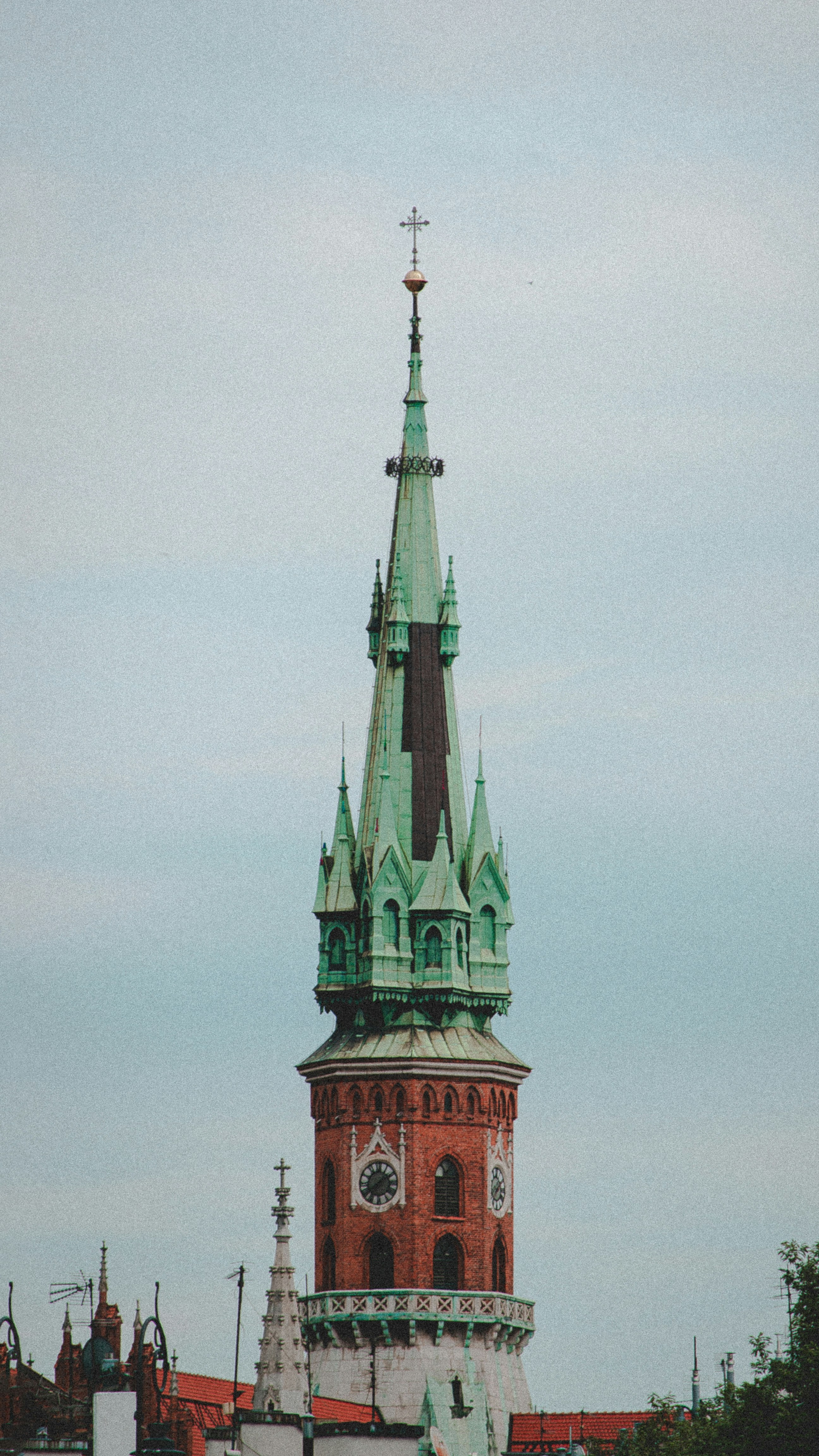 Elaborate clock tower with a green spire and red brick base, showcasing intricate architectural details against a pale sky.