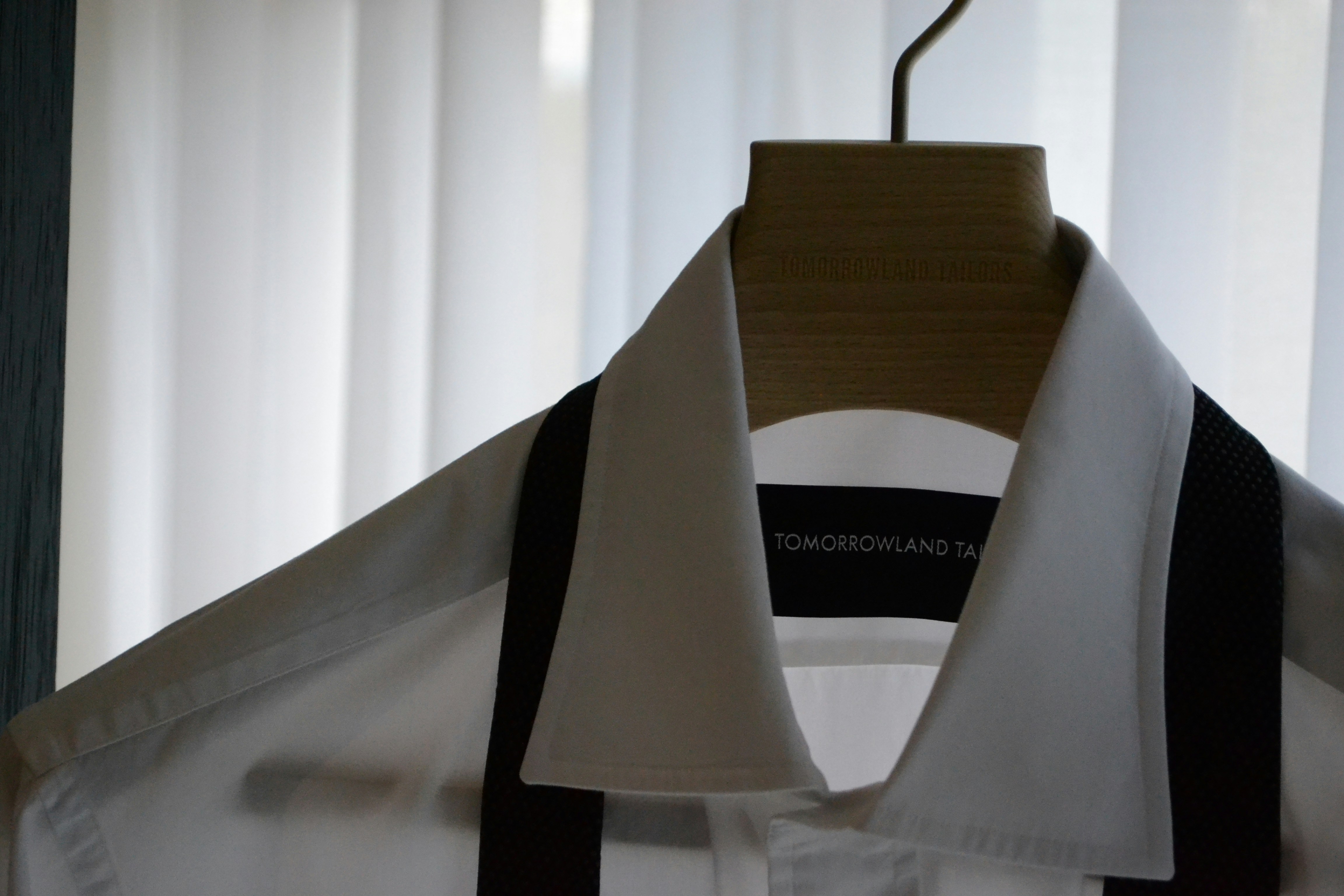 A crisp white shirt with black suspenders hangs elegantly on a wooden hanger against a backdrop of soft, diffused light from curtains.