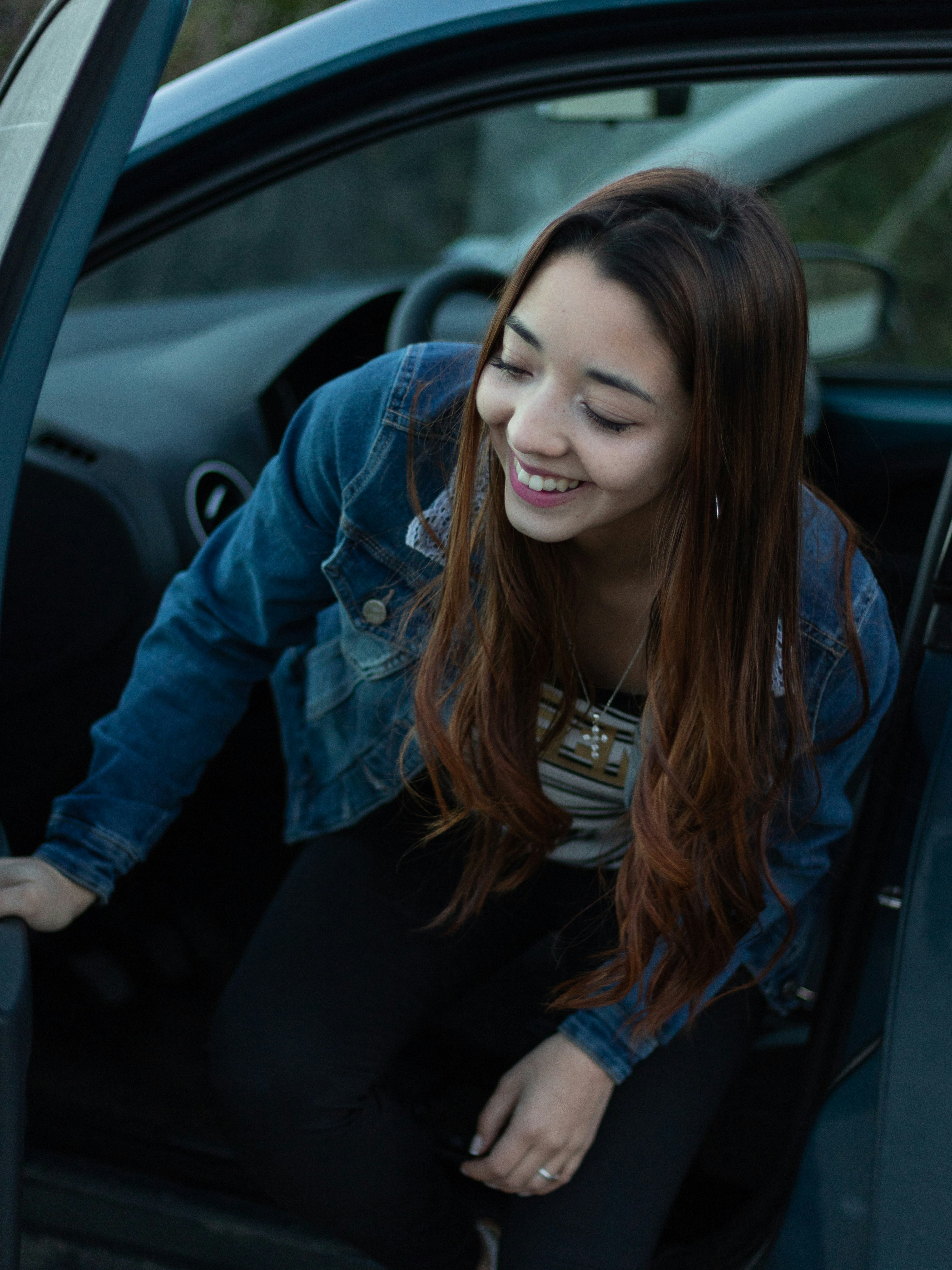 Young woman smiling as she exits a car, dressed in a denim jacket and black pants. The scene captures a light-hearted moment during a casual outing.