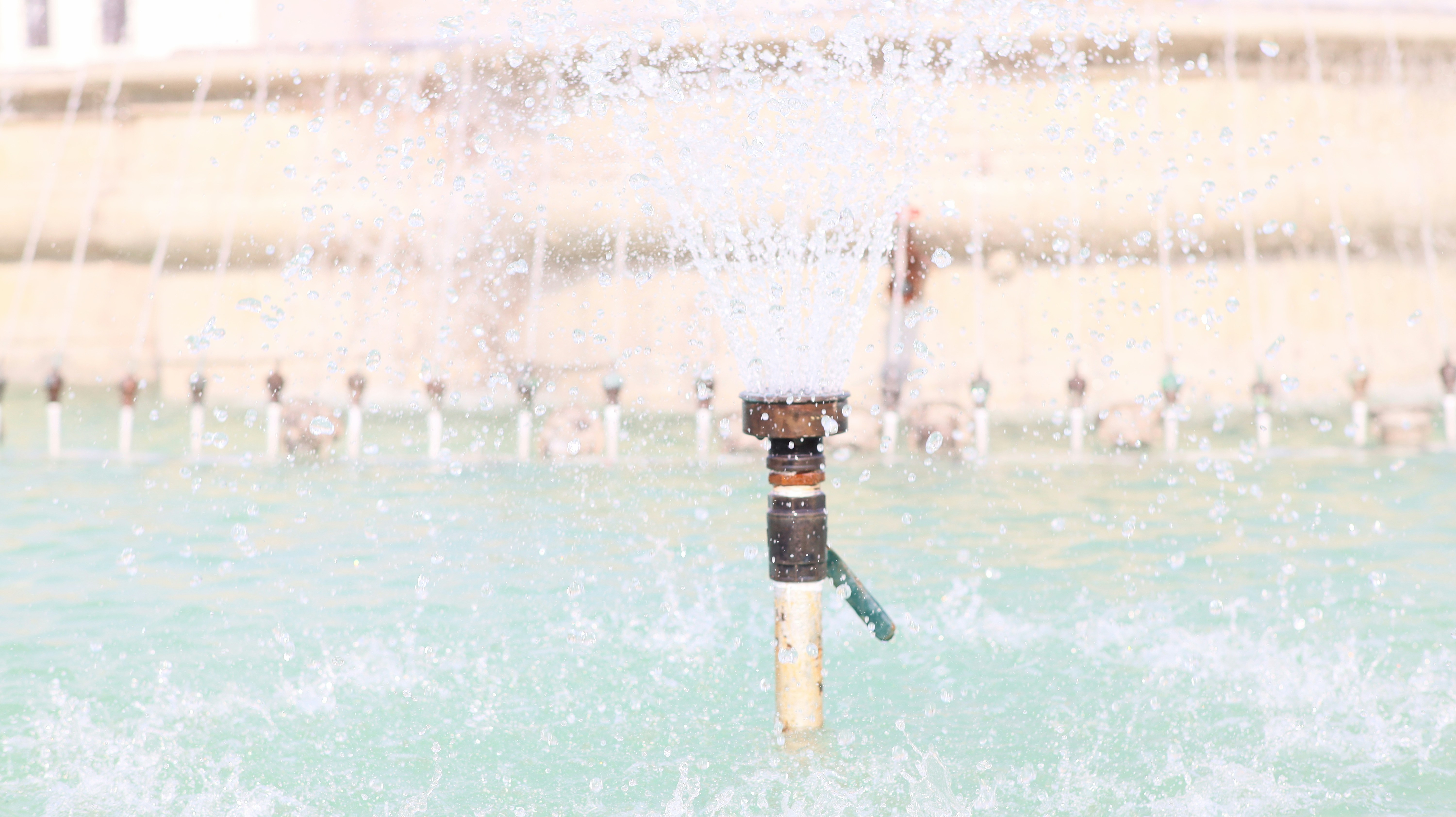 A vibrant sprinkler shoots water into the air, creating a dynamic display in a tranquil pool. The background features blurred fountain structures, enhancing the focus on the water's movement.