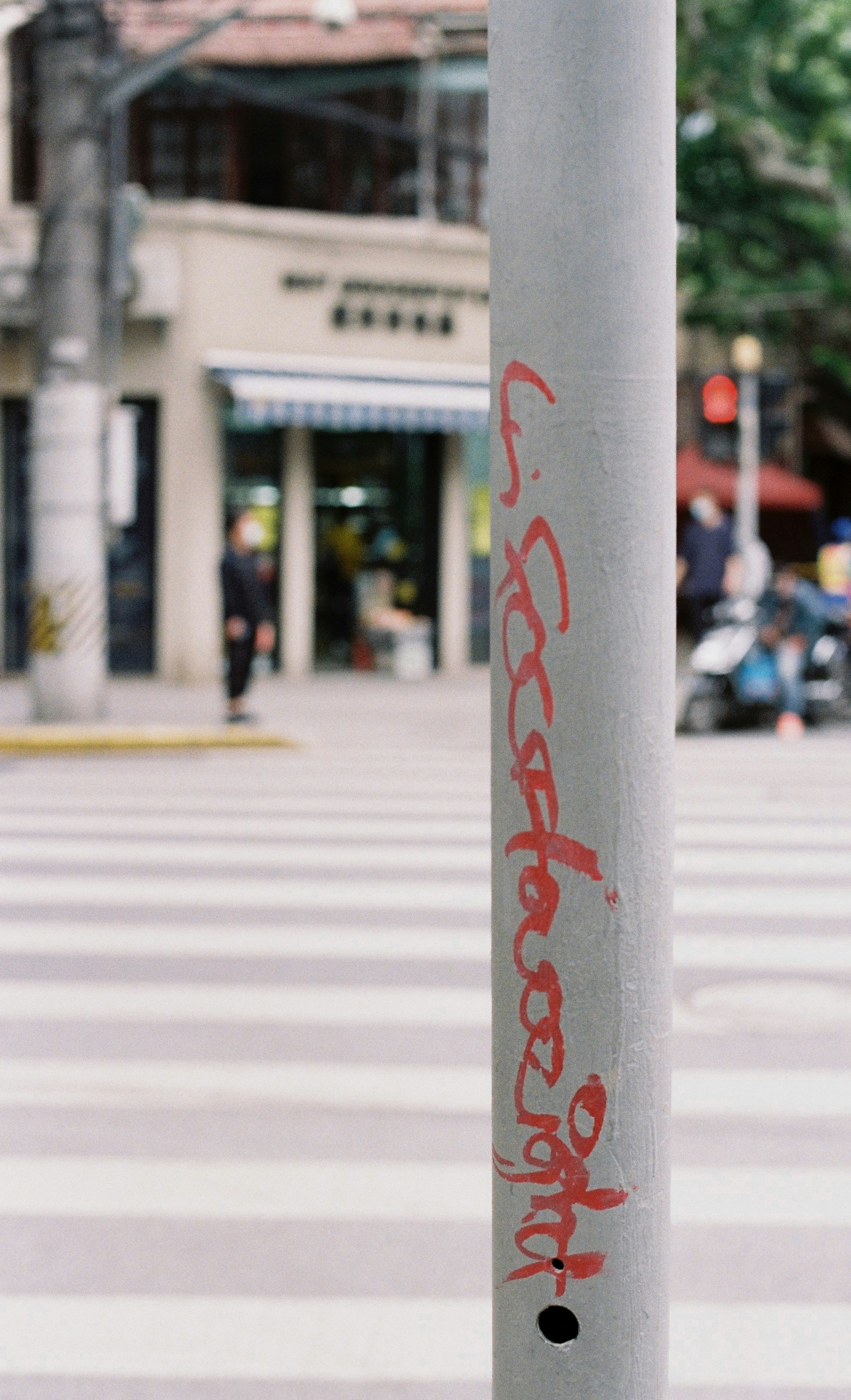 Graffiti on a pole in an urban setting, with a blurred background of pedestrians and storefronts. The vibrant red text contrasts with the muted surroundings.