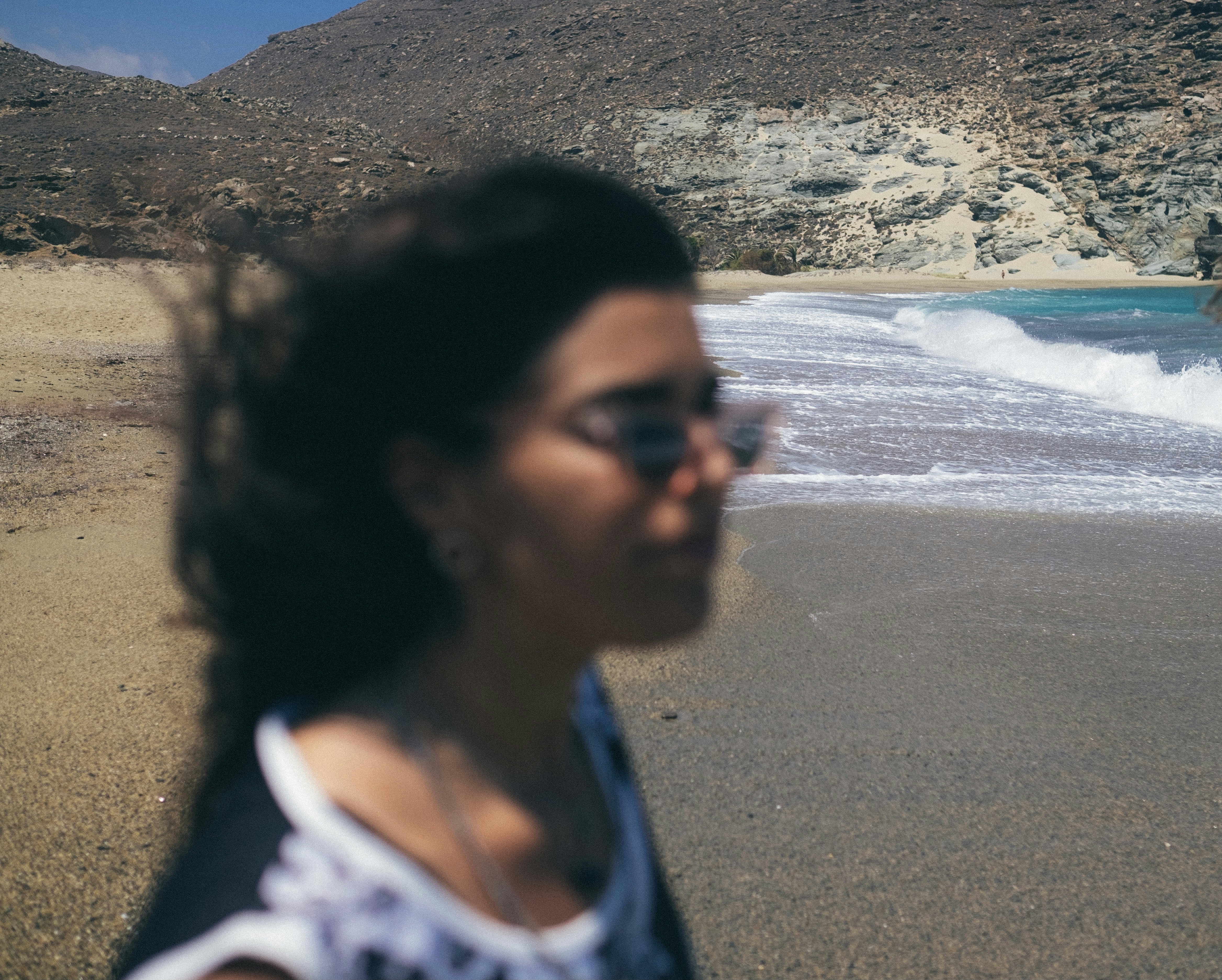 A woman stands blurred against the backdrop of a tranquil beach, where gentle waves meet the sandy shore under a clear sky.