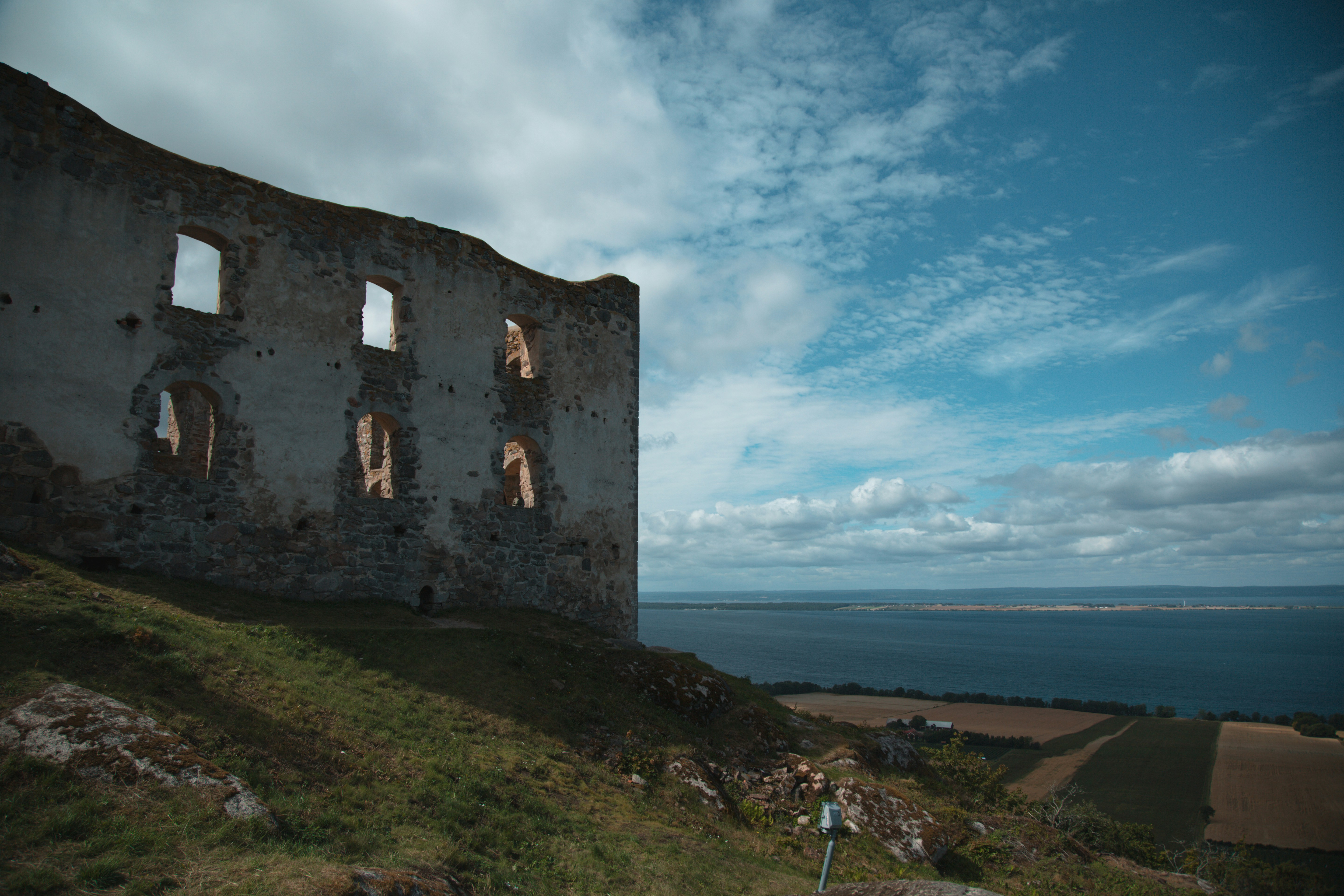 Ancient stone ruins perched on a hillside overlooking a vast ocean and farmland, capturing the essence of history against a dramatic sky.