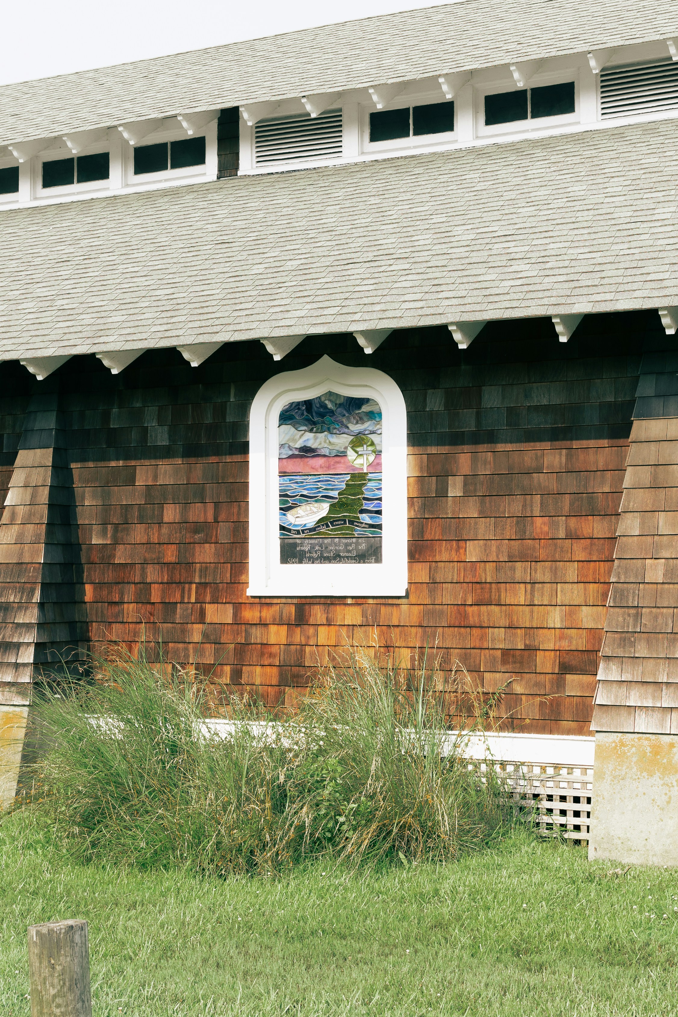 Stained glass window depicting a serene landscape framed by a wooden wall, with grass in the foreground. The artwork captures a tranquil scene of water and trees.
