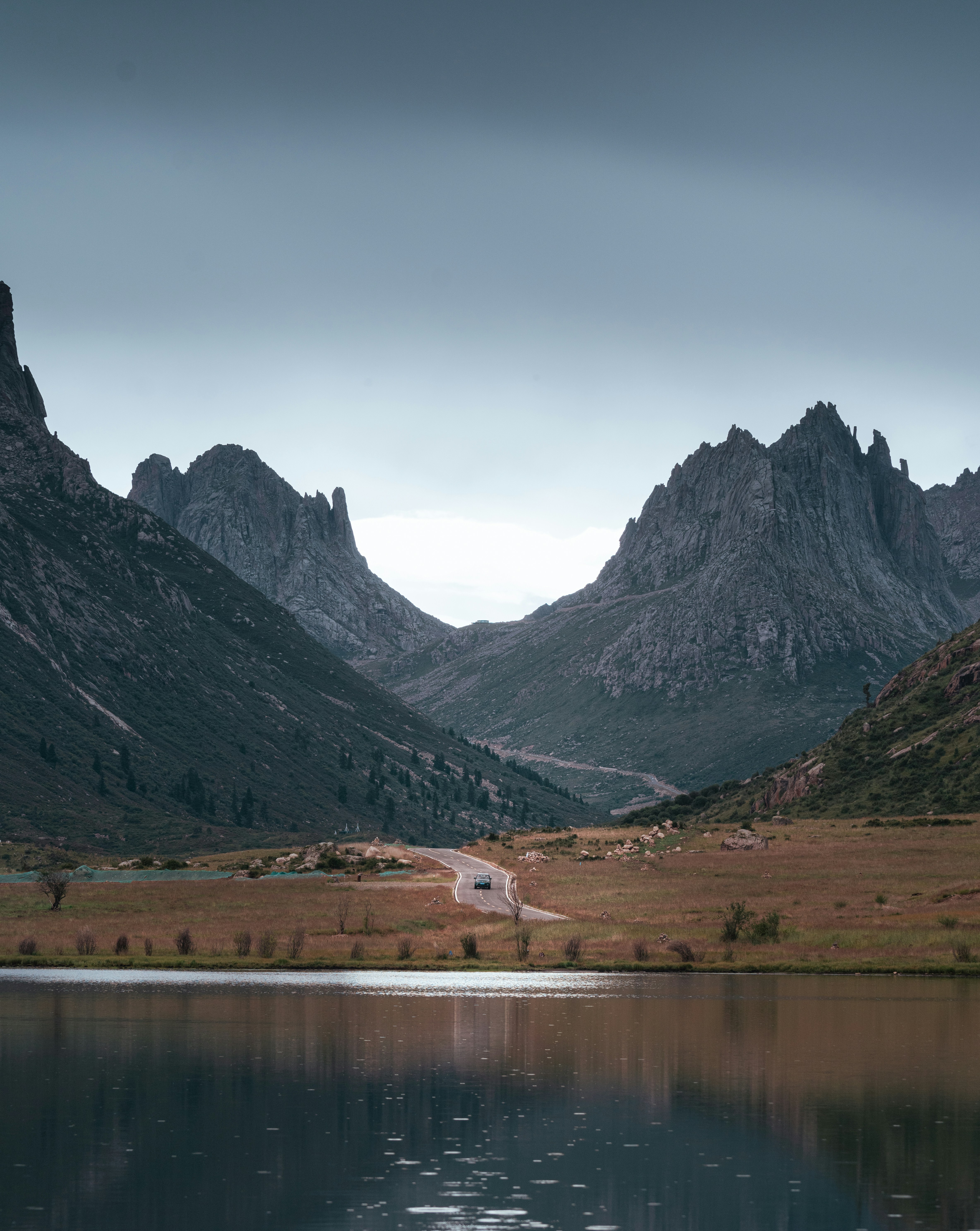 A winding road meanders through a valley flanked by towering mountain peaks under a moody sky. The reflection in the calm water adds depth to the serene landscape.